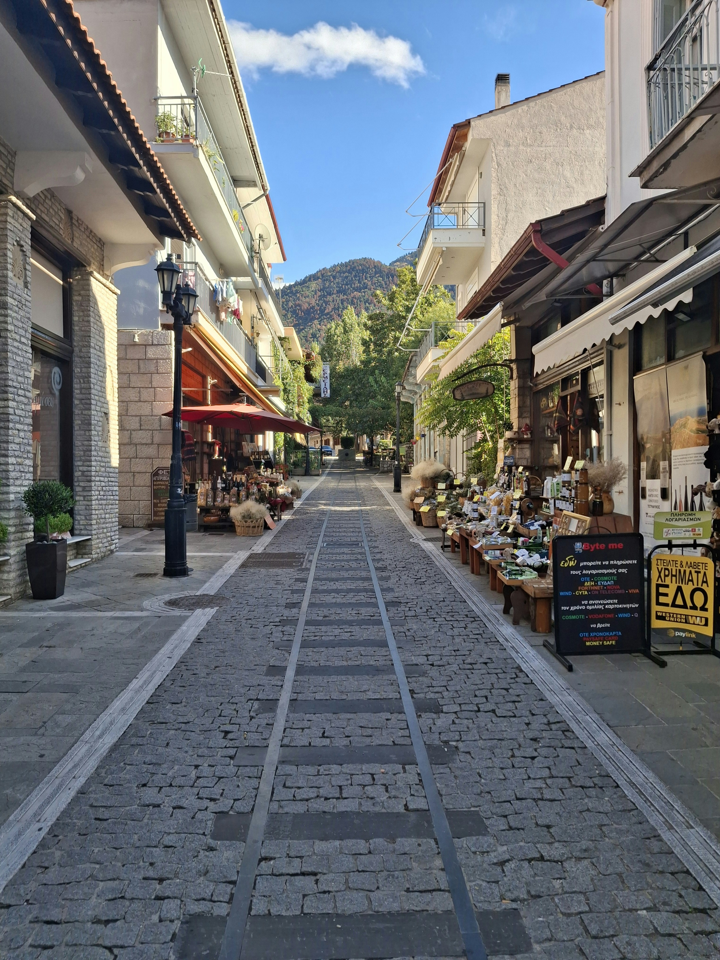 Charming street lined with shops and local produce stands, framed by lush mountains in the background. Bright blue sky enhances the vibrant atmosphere.