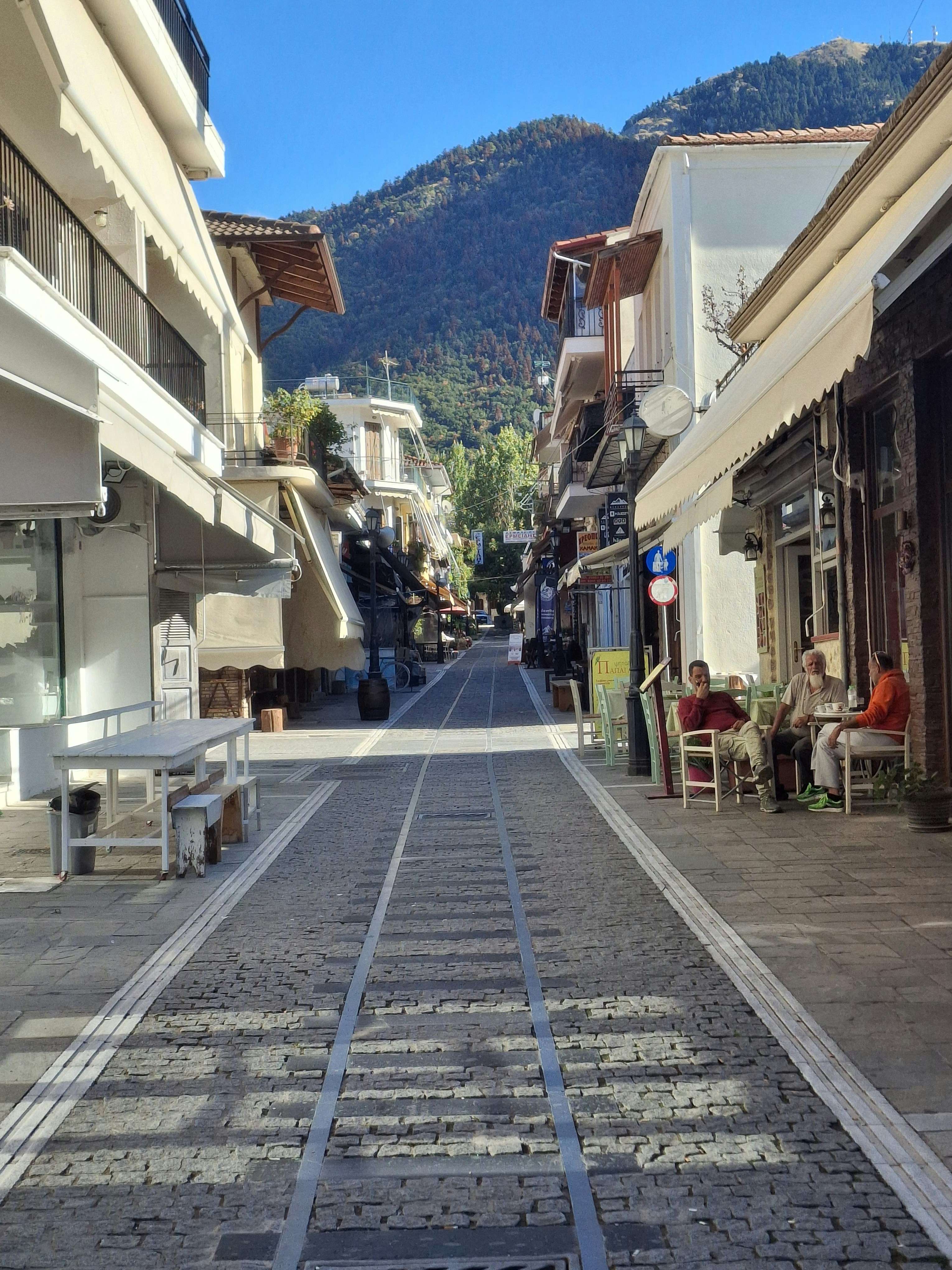 A picturesque street lined with quaint shops and cafes, framed by lush mountains in the background. The scene captures a serene moment in a vibrant town.