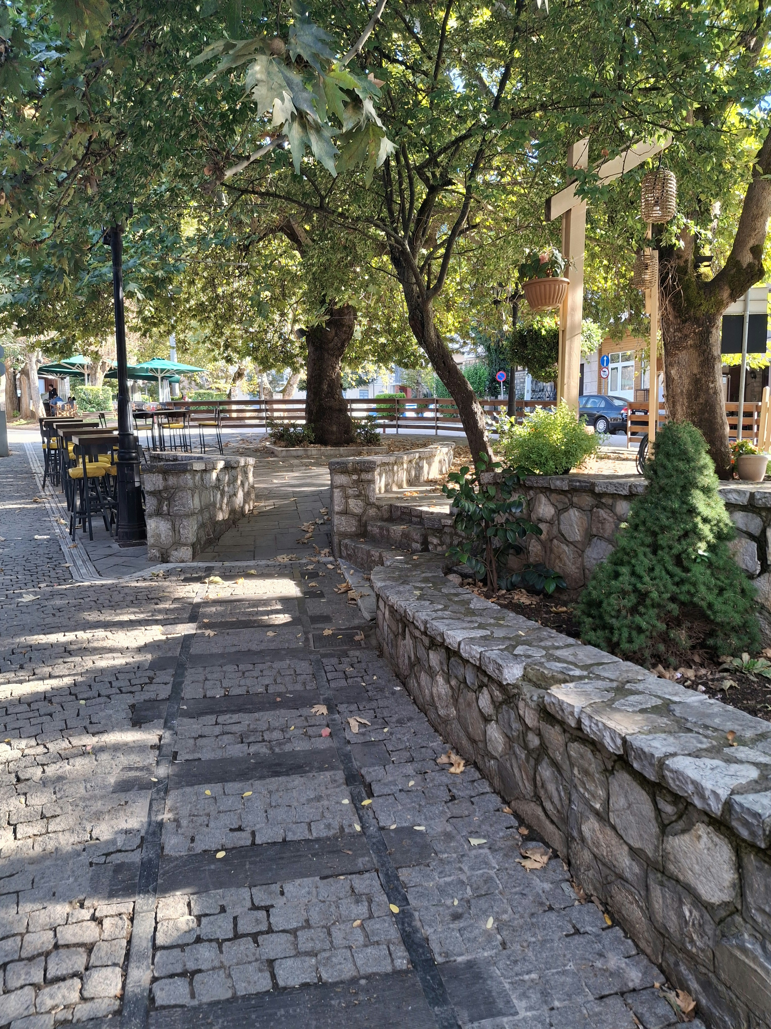 A charming cobblestone pathway bordered by lush trees and stone walls, leading to a quaint outdoor café. The scene exudes a peaceful, inviting atmosphere.