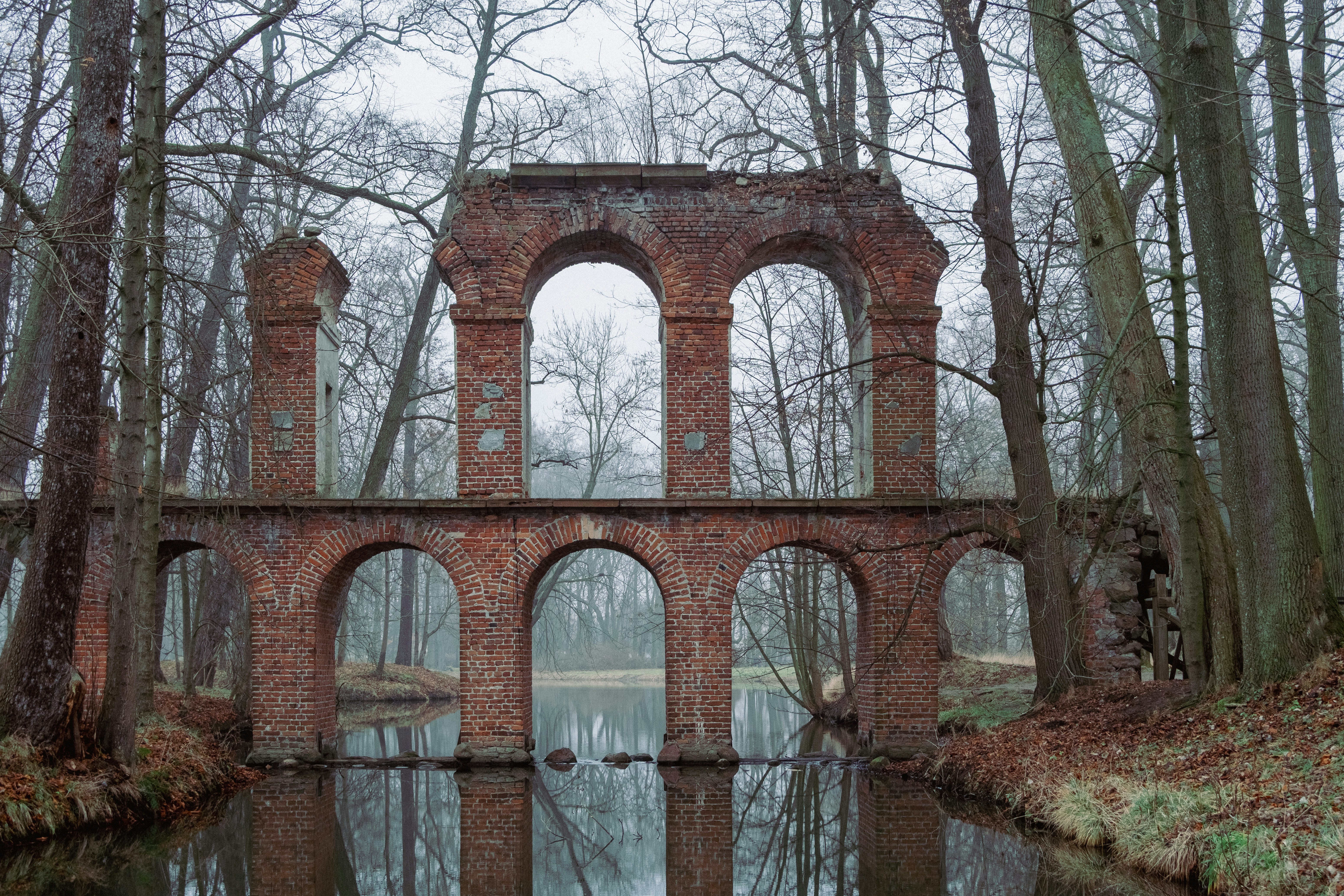 Weathered brick archway reflected in still waters, surrounded by a serene forest in misty ambiance.