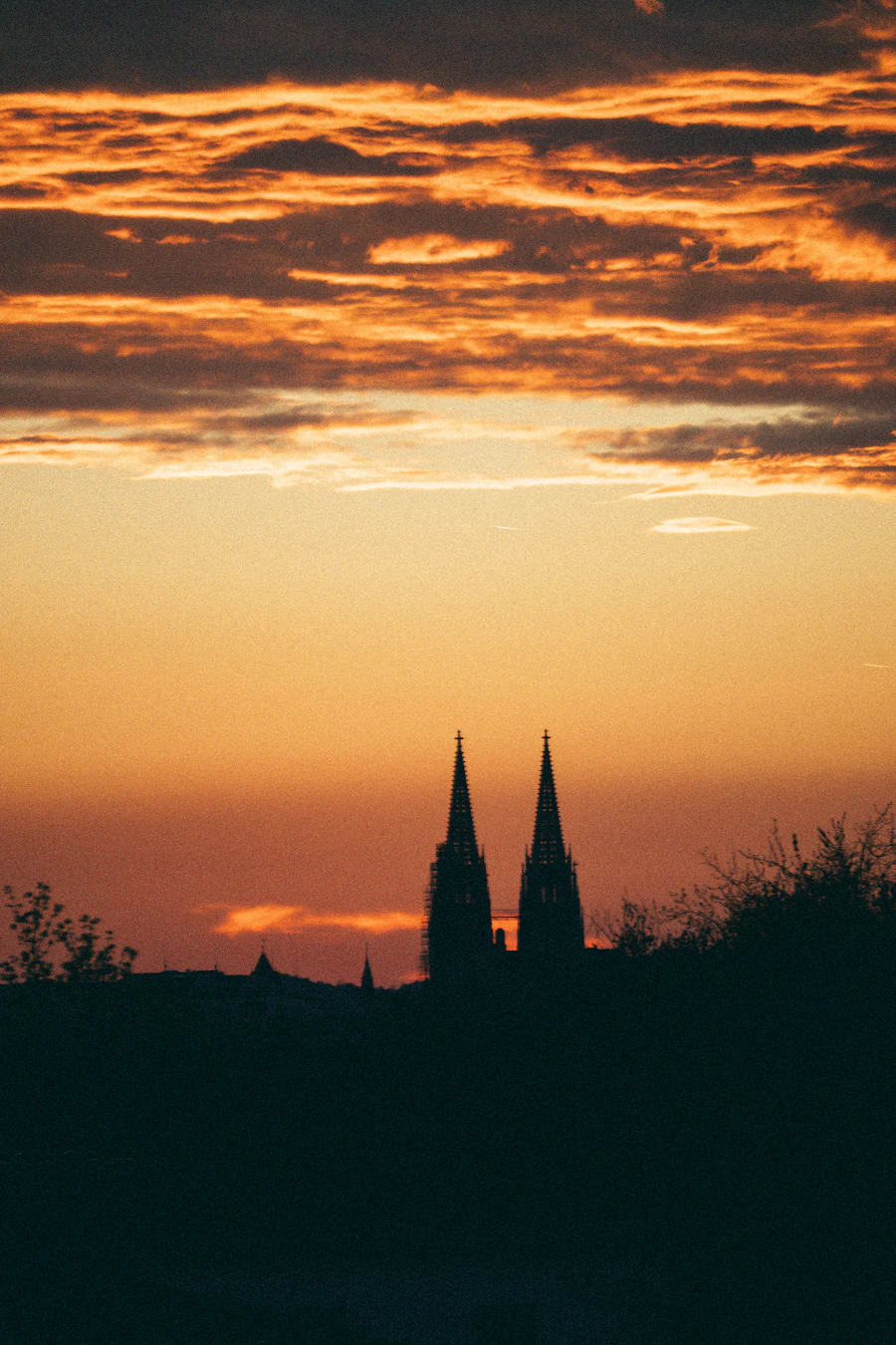 Silhouetted twin cathedral spires beneath layered orange and gold clouds at sunset.