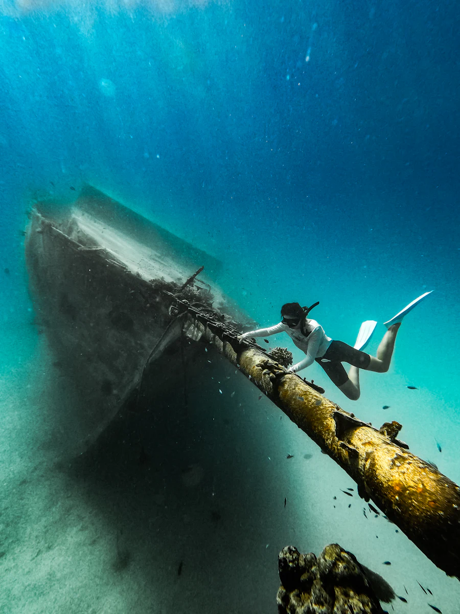 Snorkeler in fins and mask inspecting the corroded bow of a shipwreck underwater, with blue water fading into the distance.