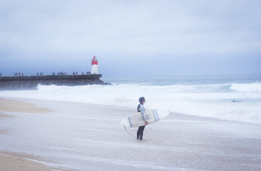 Surfer standing on the shore, holding a surfboard, with a lighthouse in the background and waves crashing nearby.