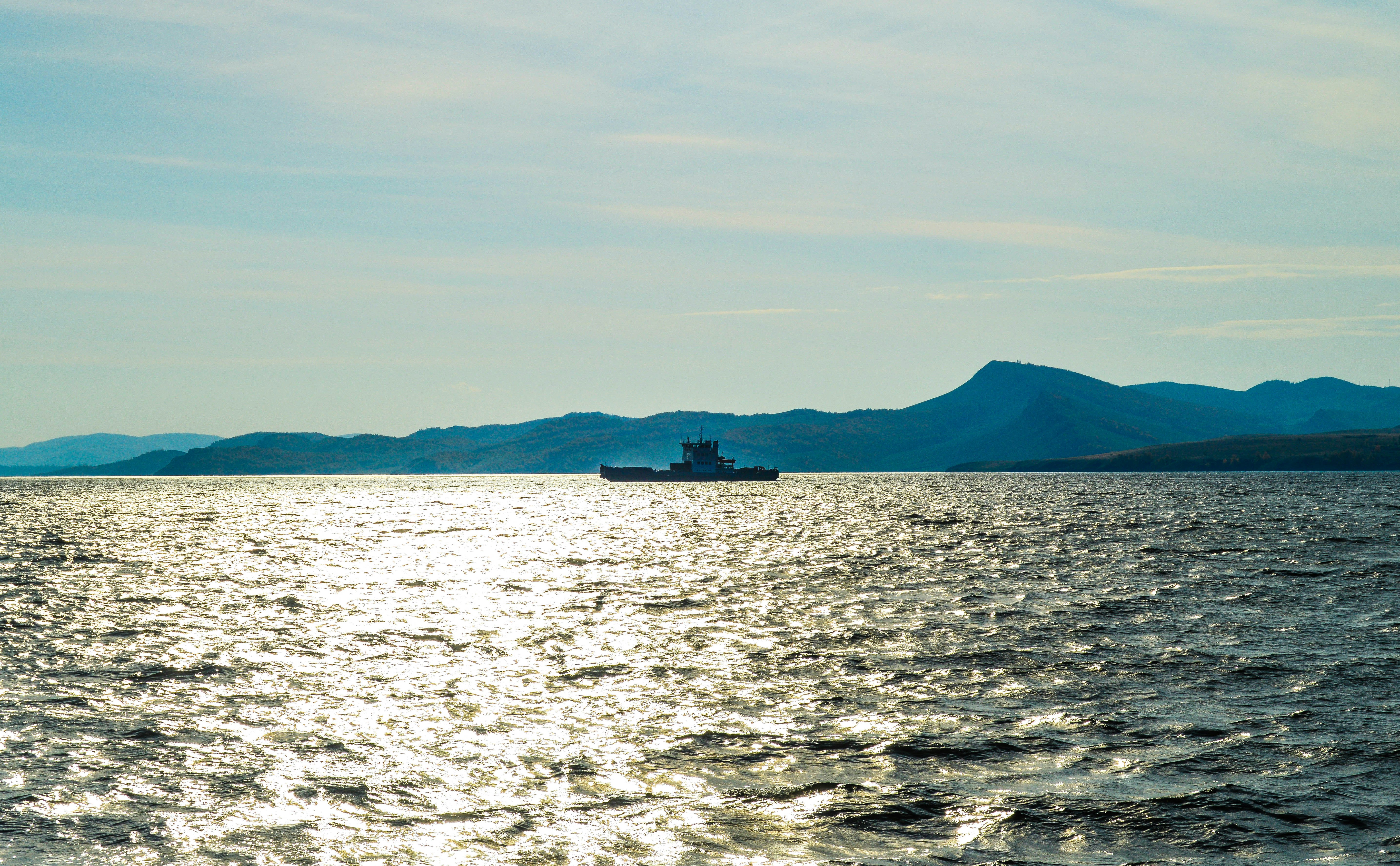 A lone ship glides across a shimmering sea, framed by distant mountains under a clear sky.