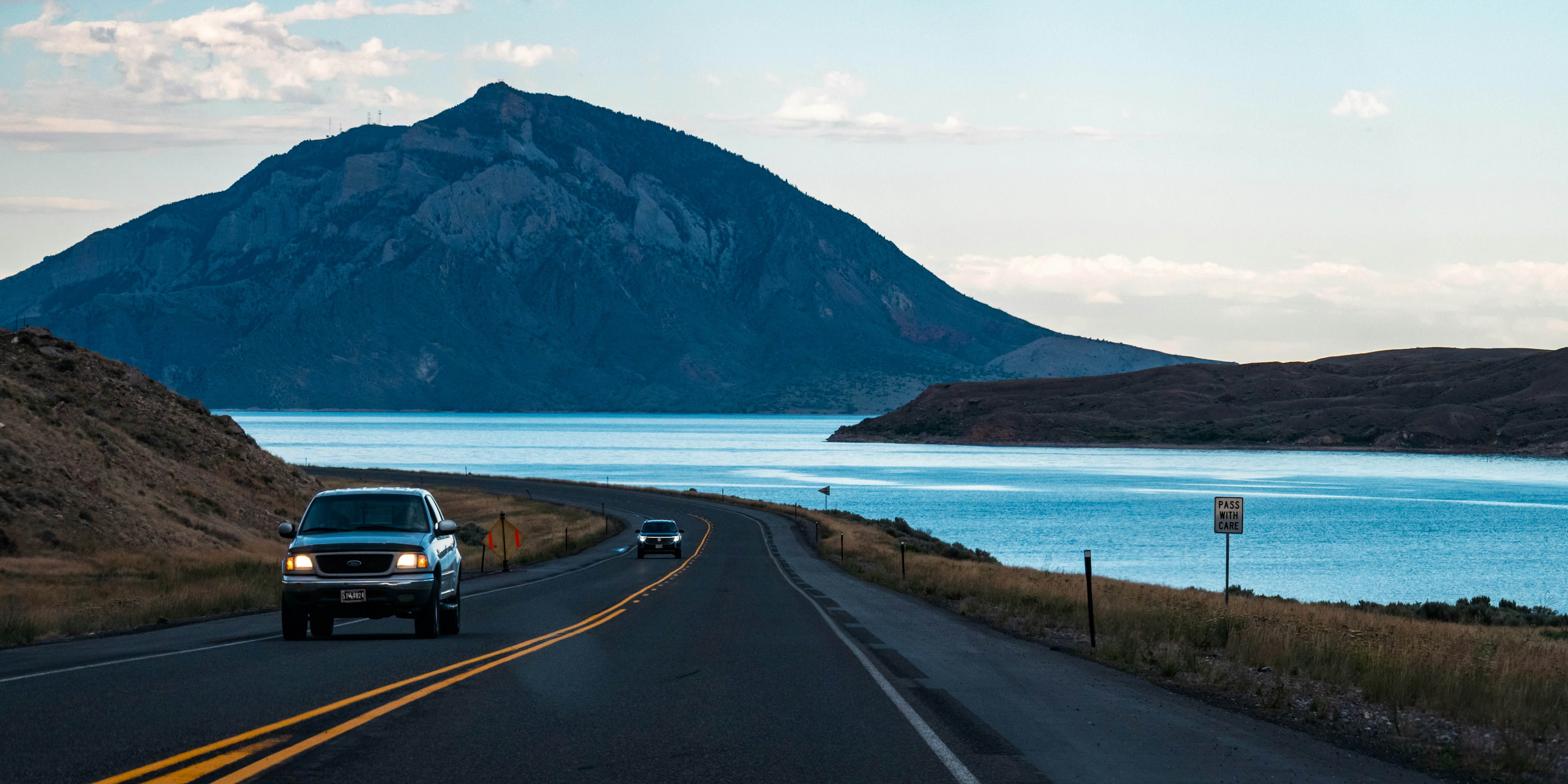 A serene road winding alongside a calm lake, with a distant mountain silhouetted against the sky. The scene captures the essence of open-road exploration.