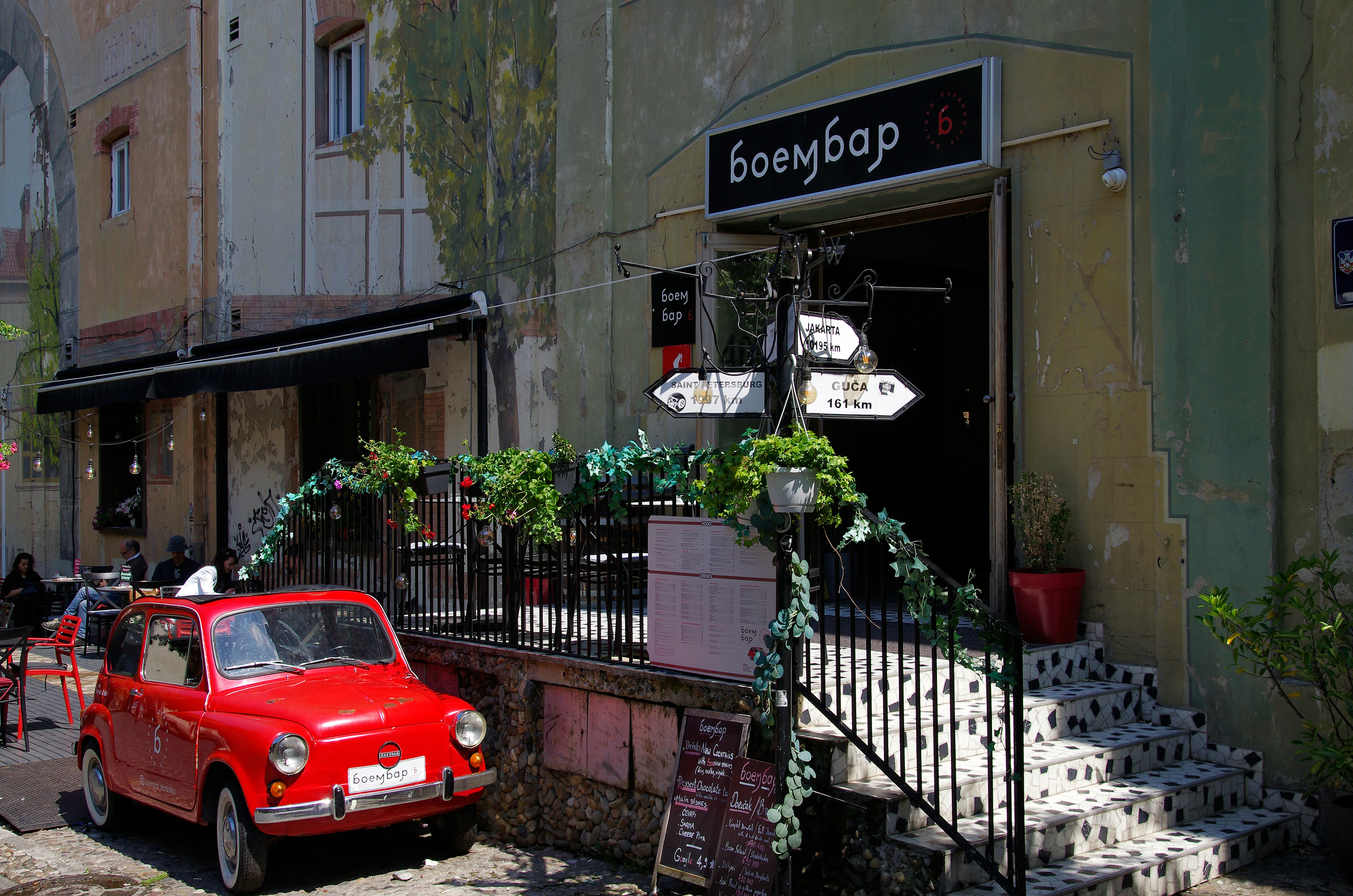 A vibrant red vintage car parked outside a cozy bar adorned with greenery and eclectic signage. The scene captures the essence of urban life and nostalgia.