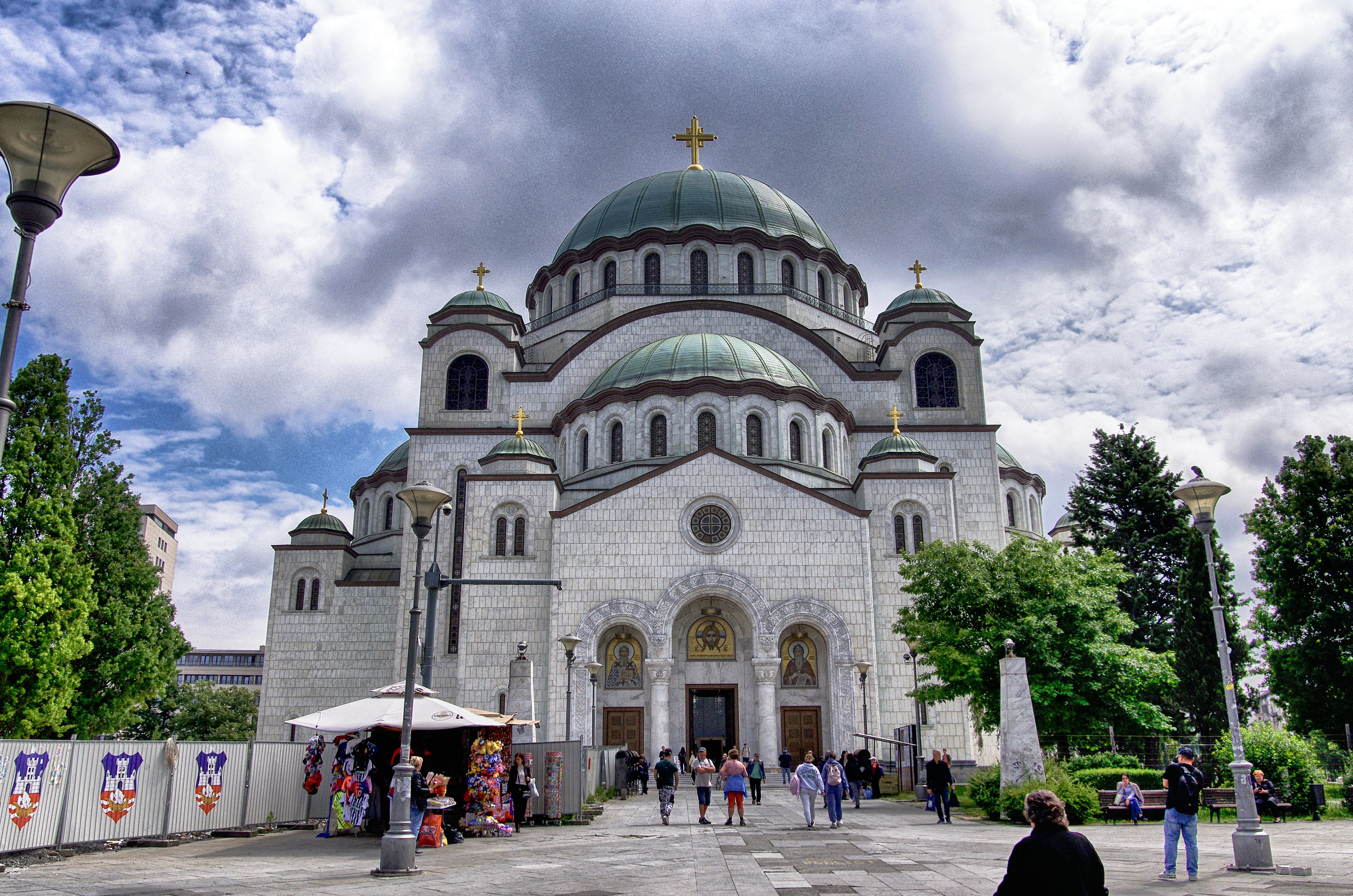 Imposing church structure with a green dome, surrounded by visitors and vibrant greenery under a dynamic sky.