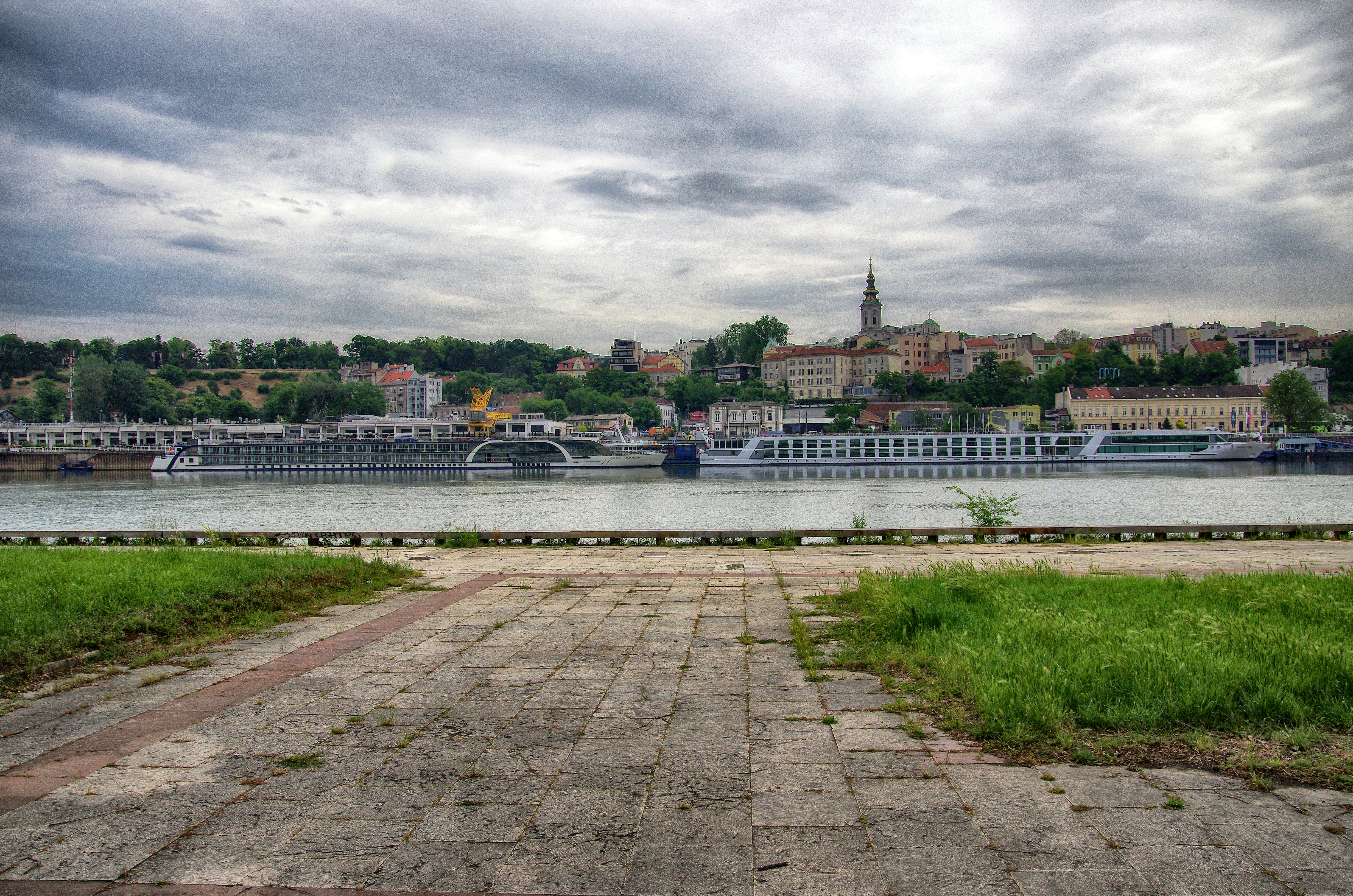 A tranquil riverside scene featuring moored boats along the bank, with a backdrop of colorful buildings and a clock tower under a cloudy sky.