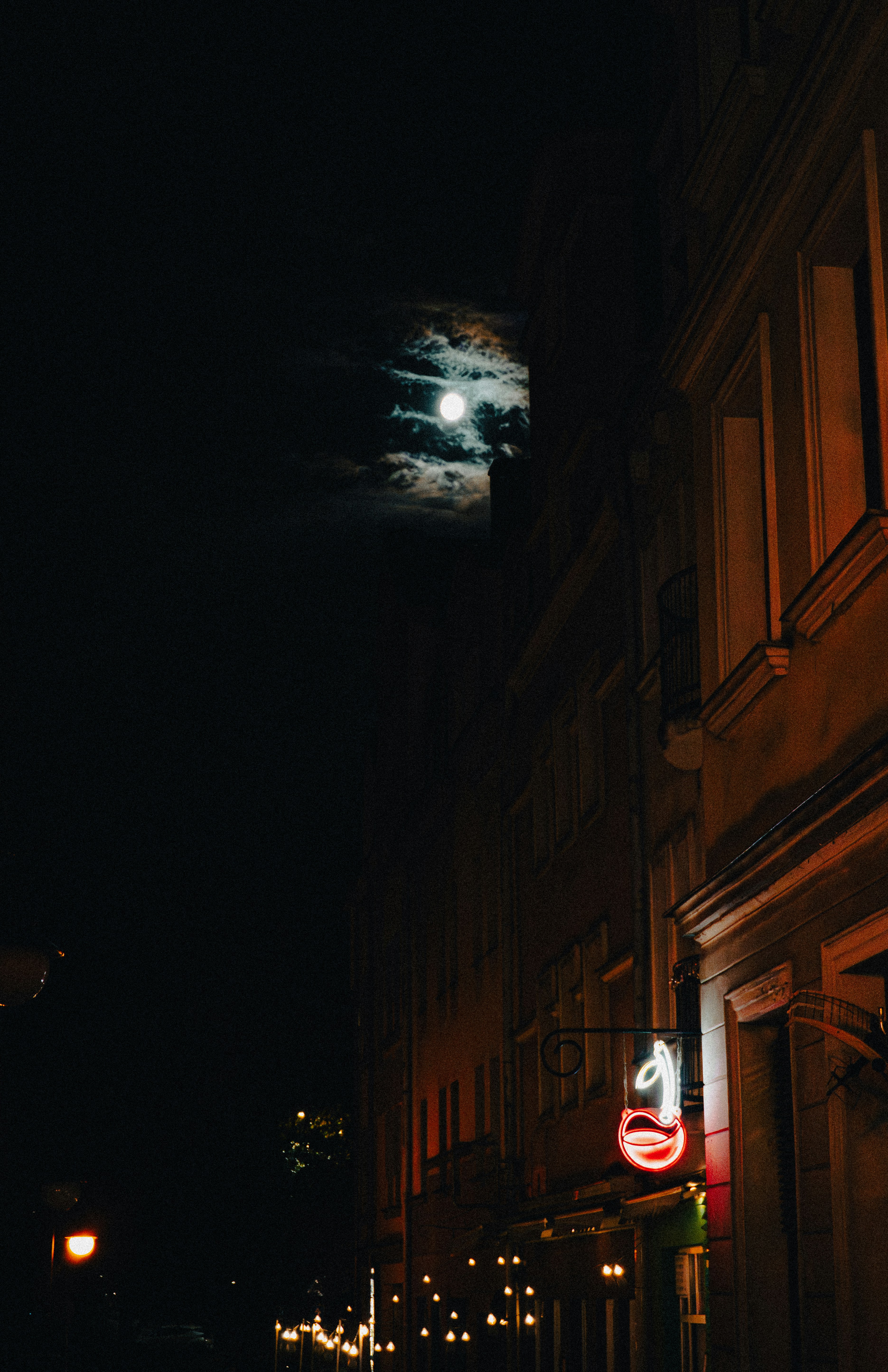 A glowing full moon peeks through wispy clouds above a dimly lit street, where a neon sign adds a splash of color to the night. The atmosphere is serene yet vibrant.
