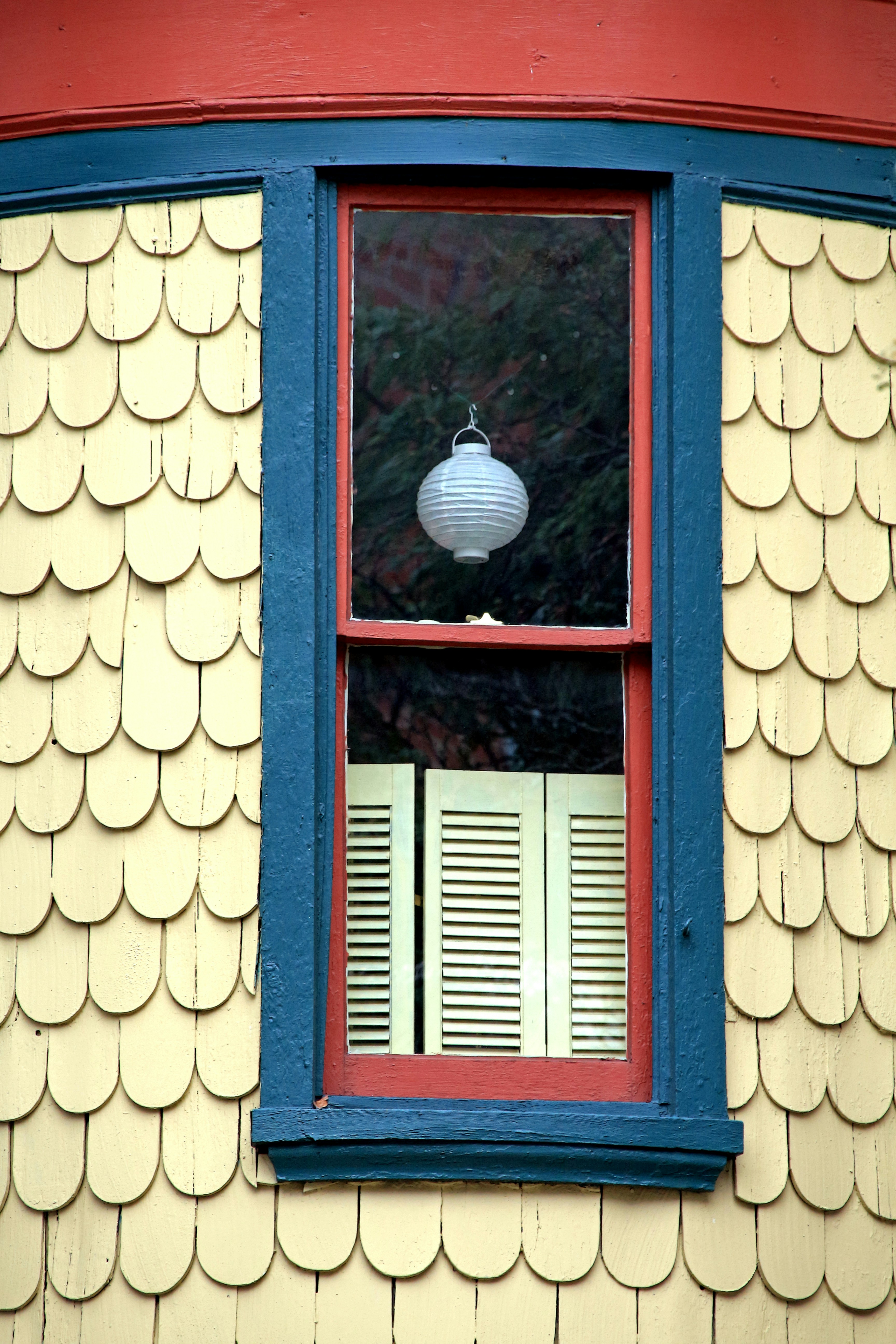 A charming window adorned with a hanging lantern, framed by vibrant blue and yellow shingles, revealing a glimpse of shuttered interiors.