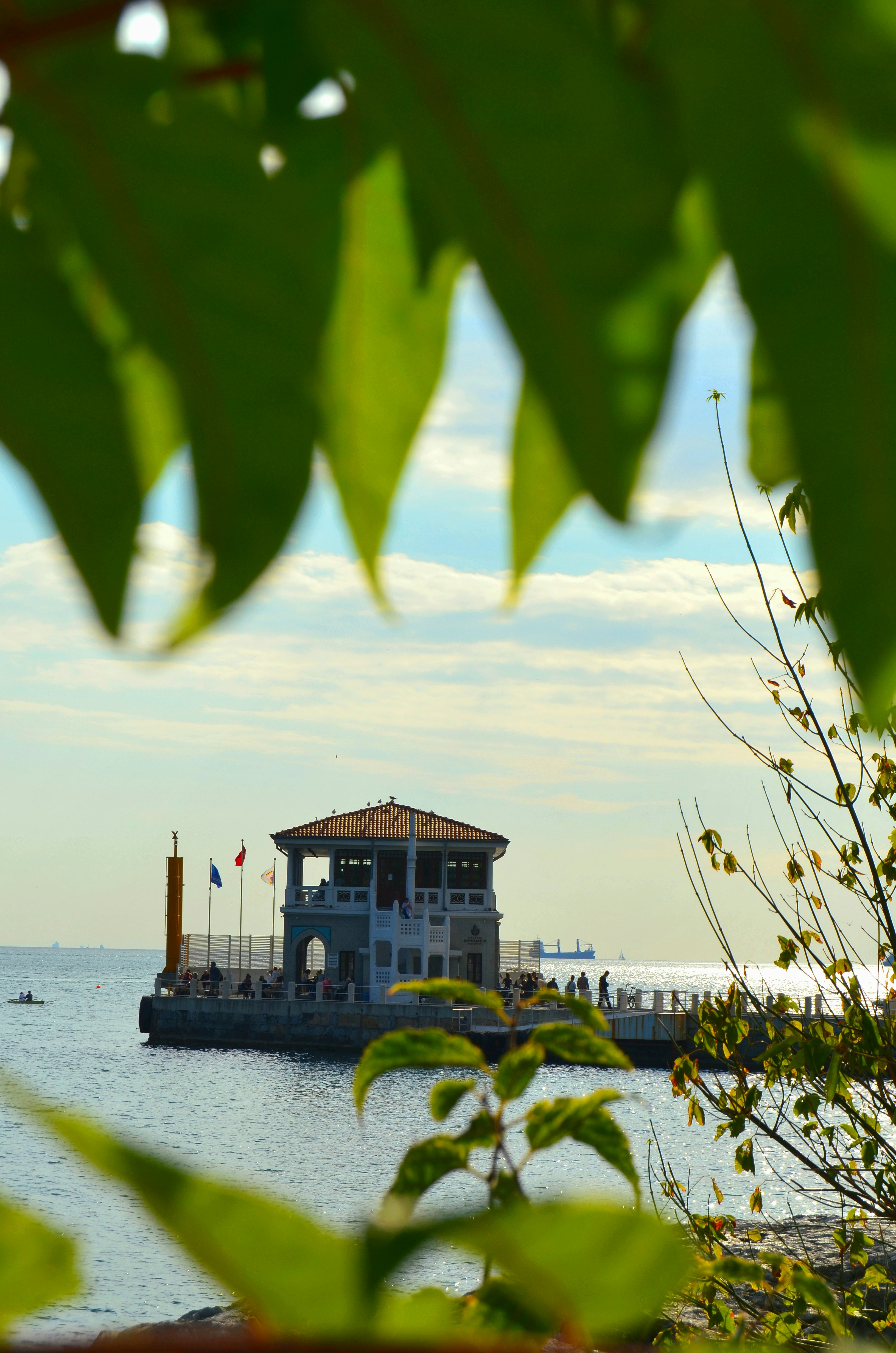 A charming waterfront structure framed by lush green leaves, with flags fluttering in the breeze. The serene water reflects the soft light of the sky.
