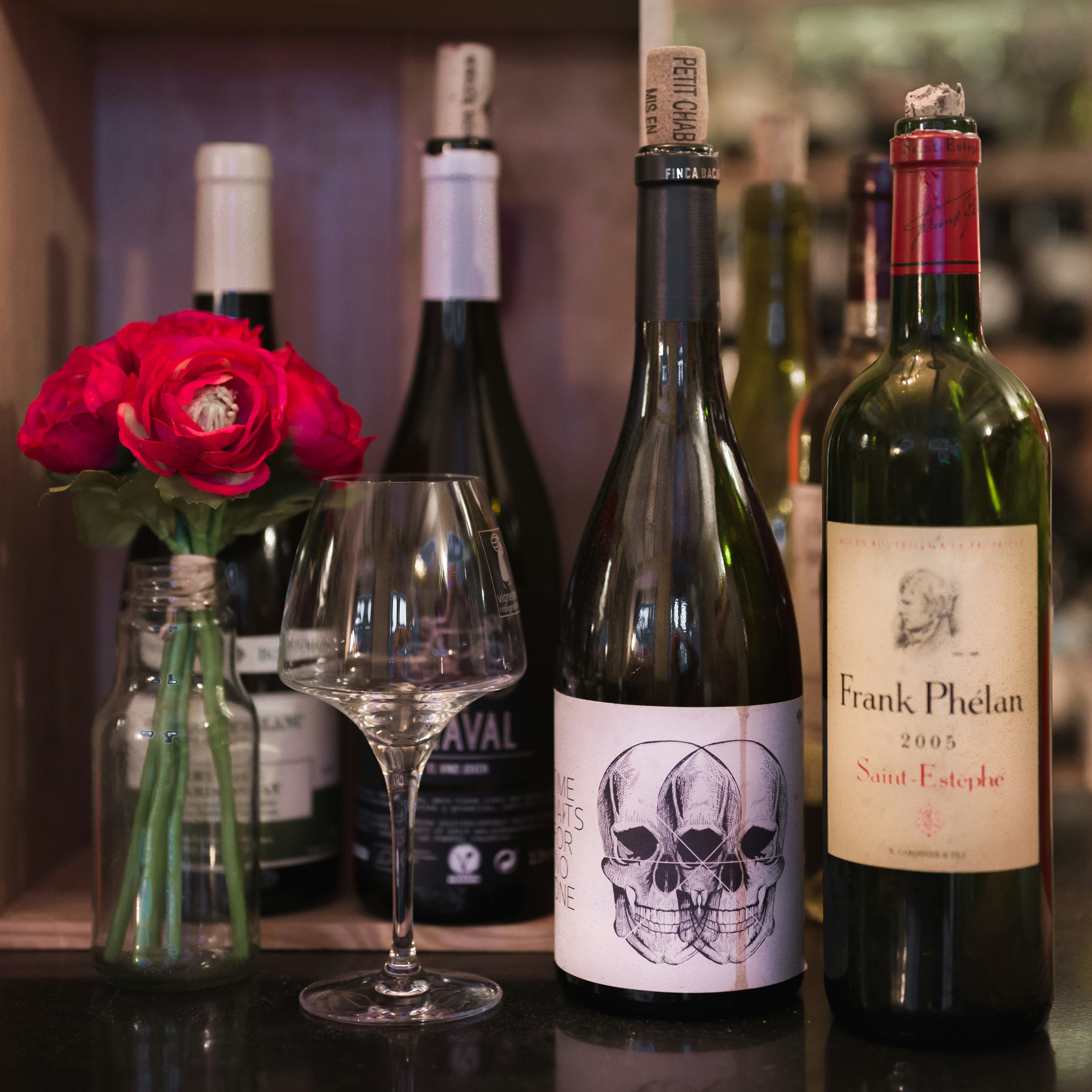 A cozy, close-up still life on a bar or wooden shelf: several wine bottles, an empty stemmed wine glass, and a small glass vase holding a bright red flower. In the foreground are two prominent bottles—one with a modern label featuring overlapping skull illustrations, and another green Bordeaux-style bottle labeled “Frank Phélan 2005 Saint-Estèphe.” Additional bottles with corks and white caps sit behind them, softly out of focus. Warm lighting and reflections on the glass give the scene an intimate wine-bar or cellar vibe. Restaurant "La Bouteillerie", Lyon, France. 2020. | Wine bottles, glass, and red flowers on counter