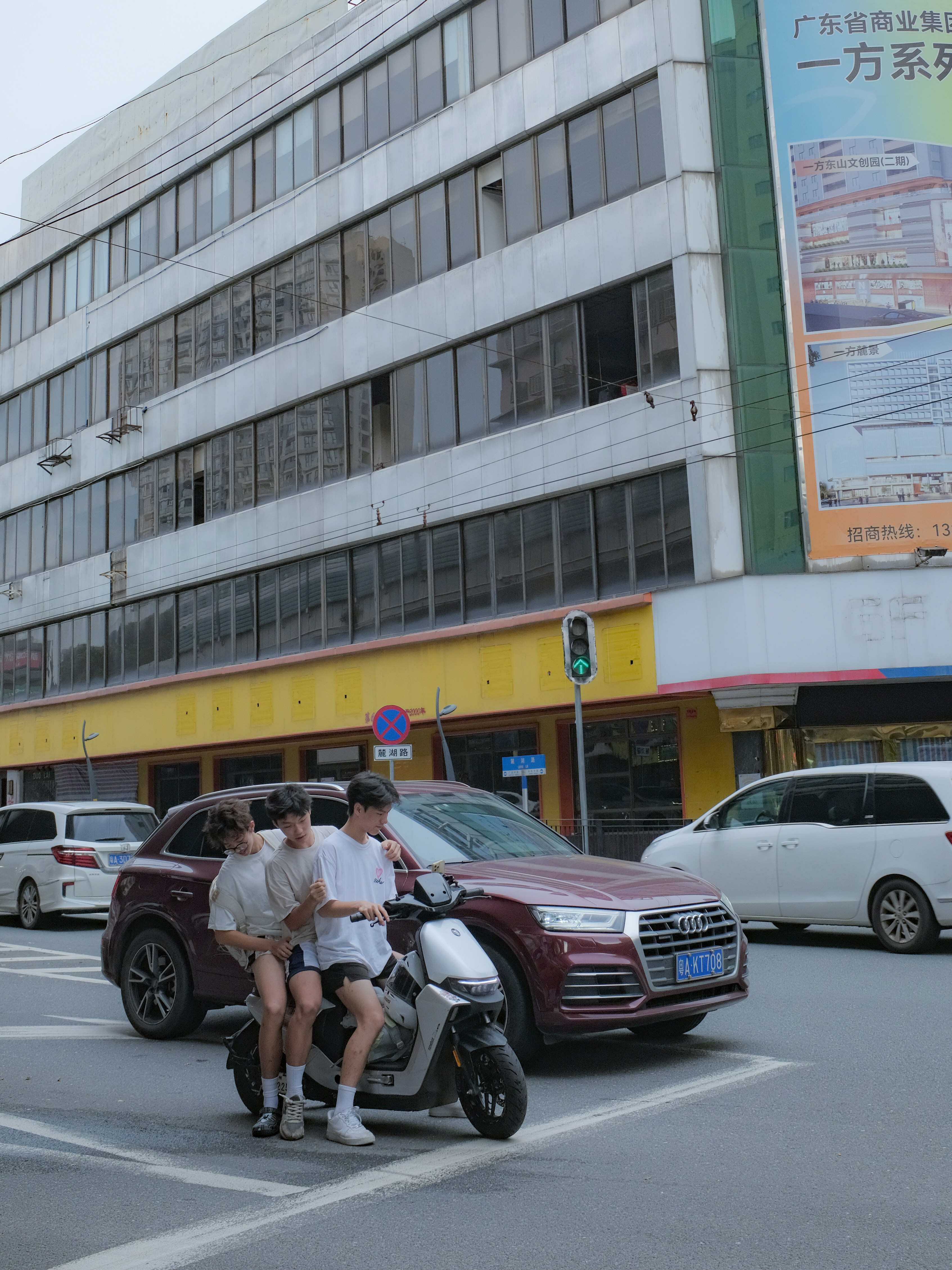 Two young men sharing a scooter at a busy intersection, surrounded by urban architecture and vehicles. The scene captures a blend of city life and camaraderie.