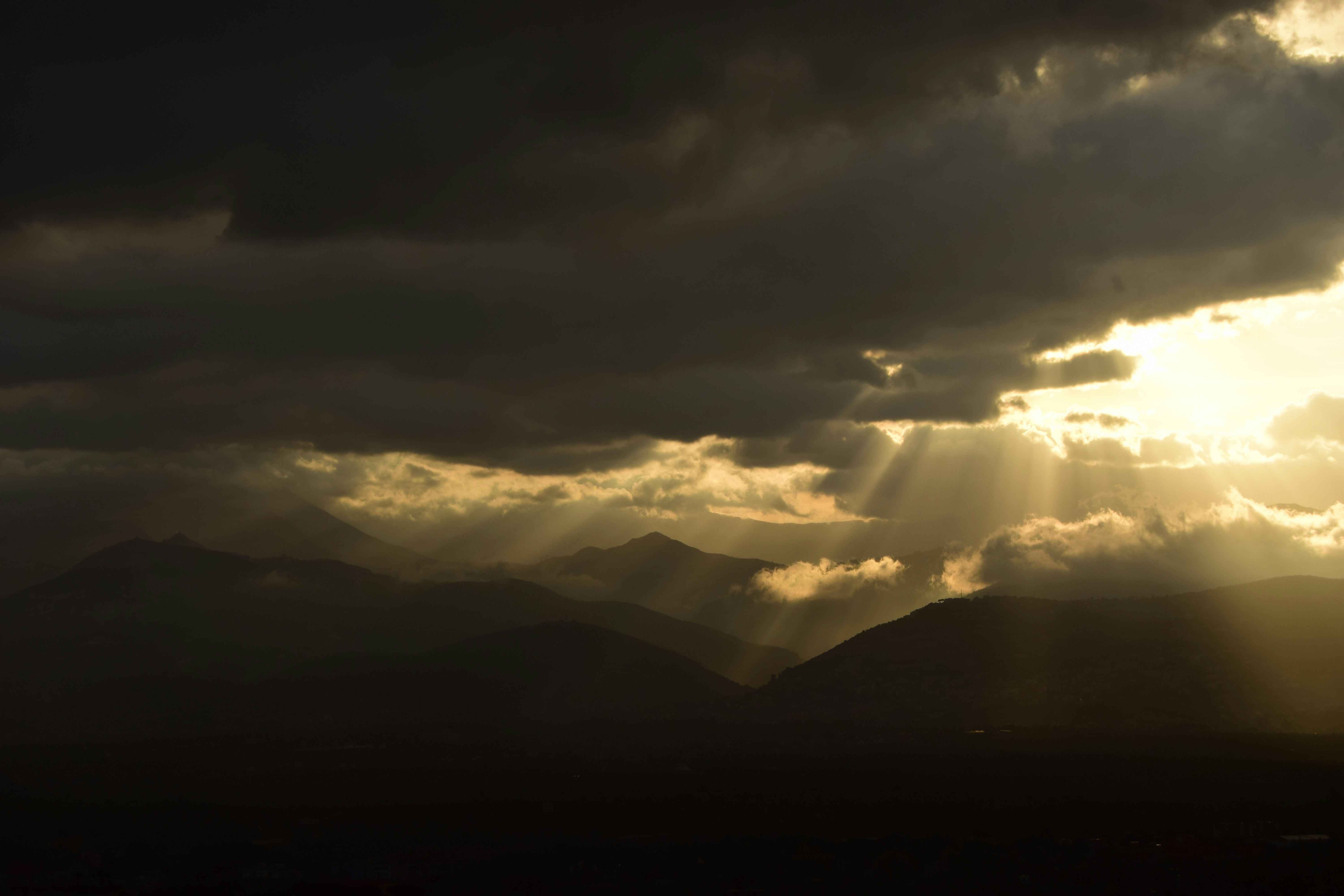 Golden rays of sunlight break through dark clouds, illuminating distant mountain ranges in a dramatic landscape.