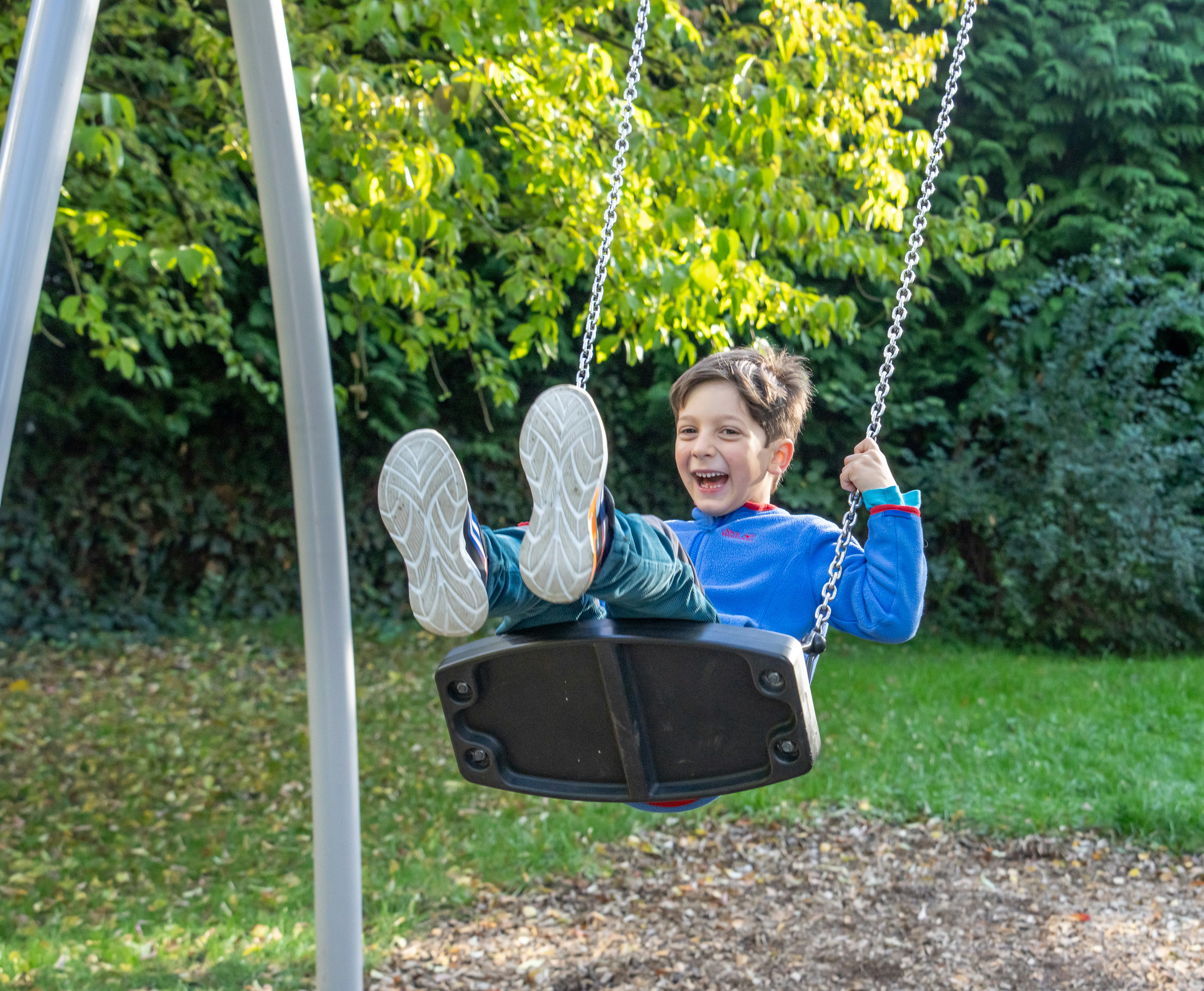 A young boy joyfully swinging on a playground swing, surrounded by lush greenery.