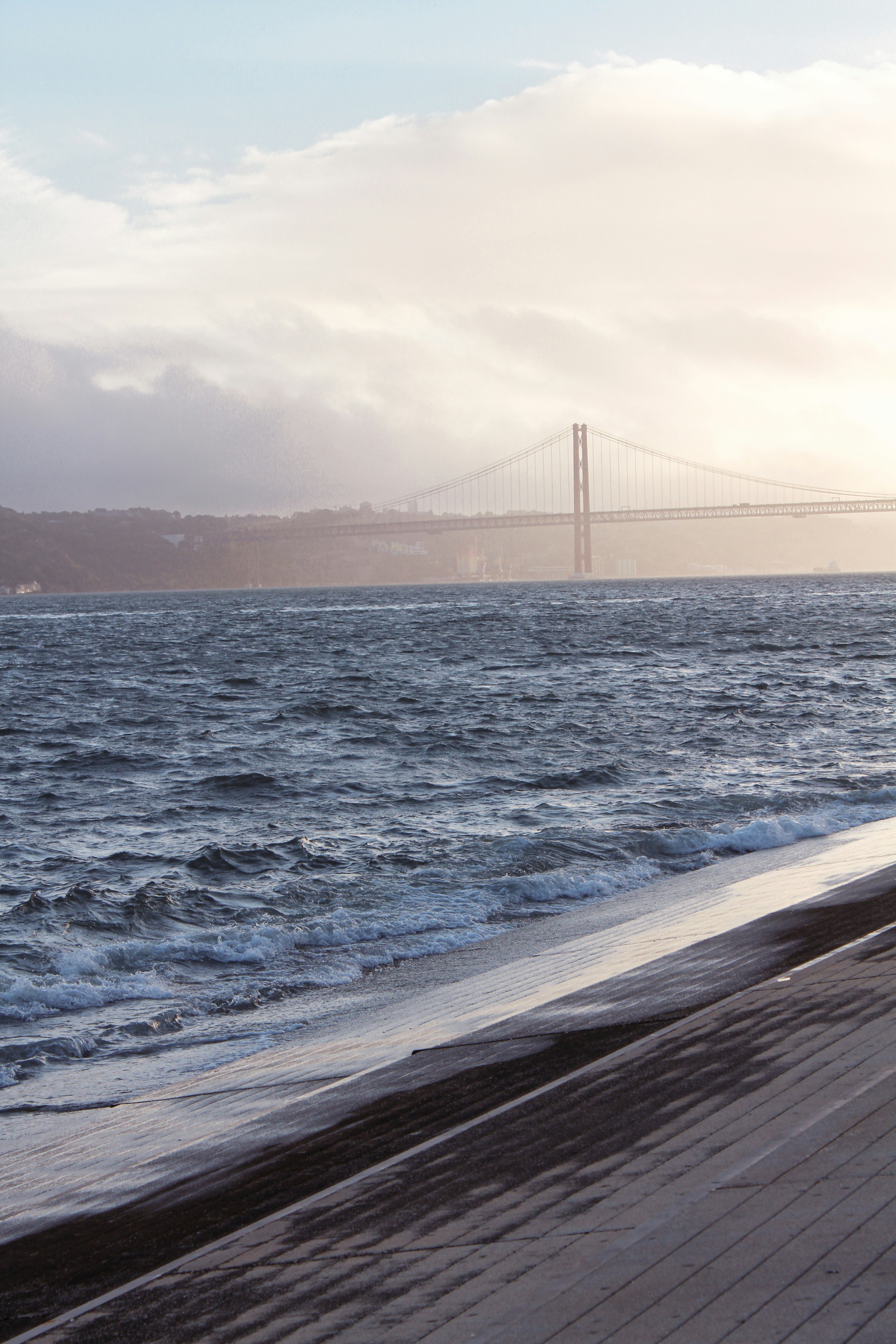 Waves gently lap against a sandy shore, with a distant bridge silhouetted against a hazy sky. The scene conveys tranquility and the passage of time.