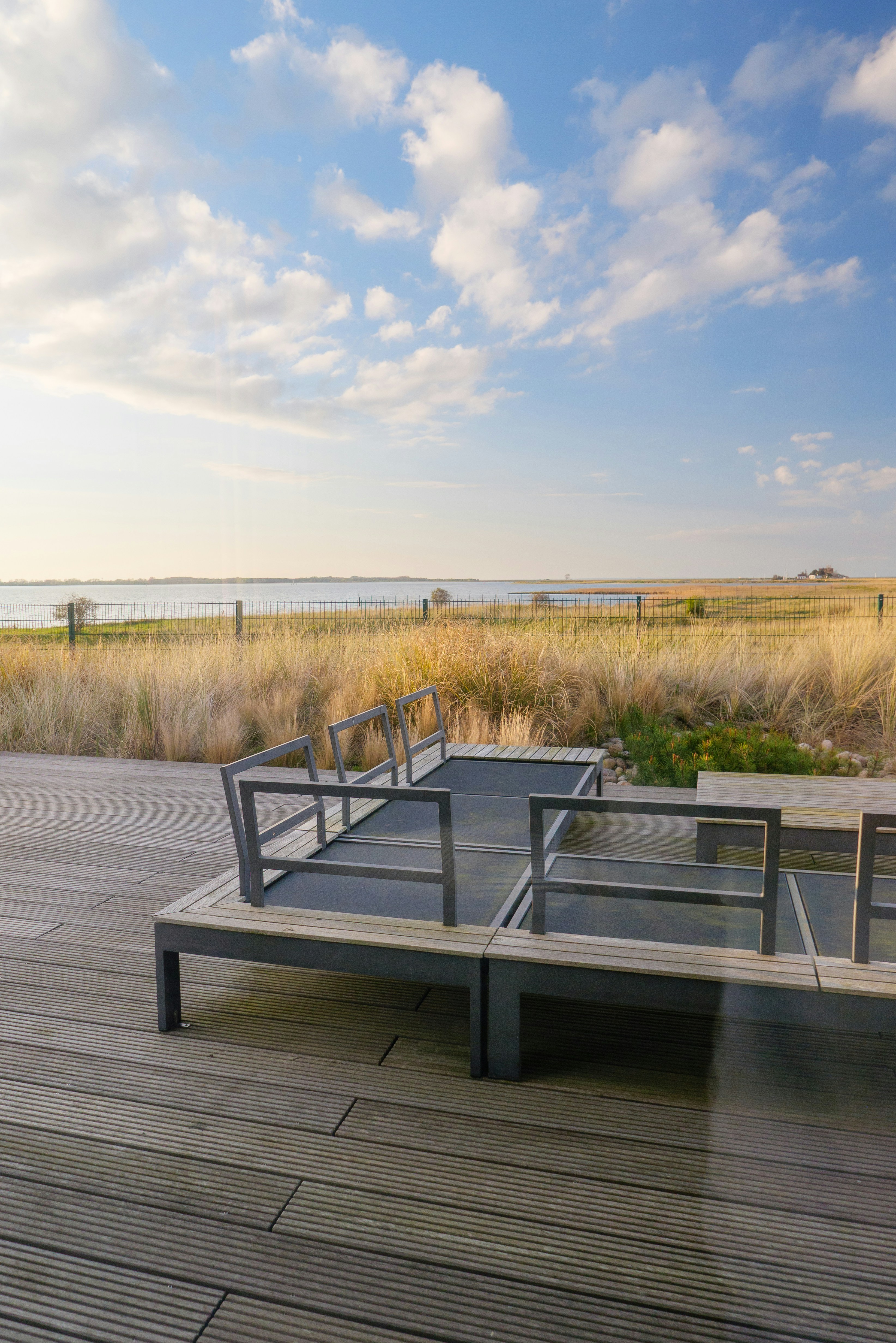 Contemporary outdoor seating area overlooking a tranquil waterway, framed by tall grasses under a clear blue sky.