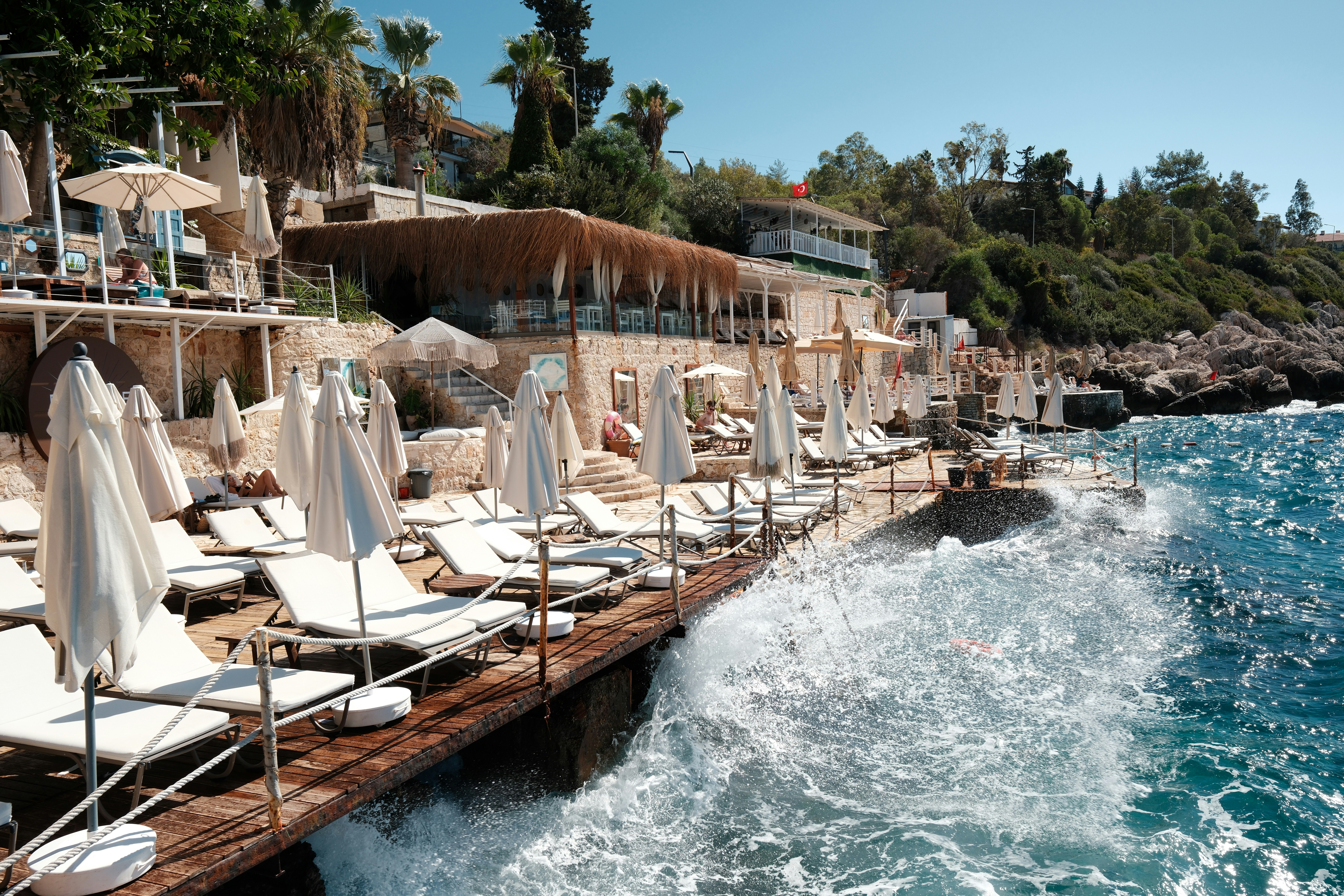 Lounge chairs lined along a waterfront deck, with umbrellas providing shade as waves crash nearby. The scene captures a tranquil coastal retreat.