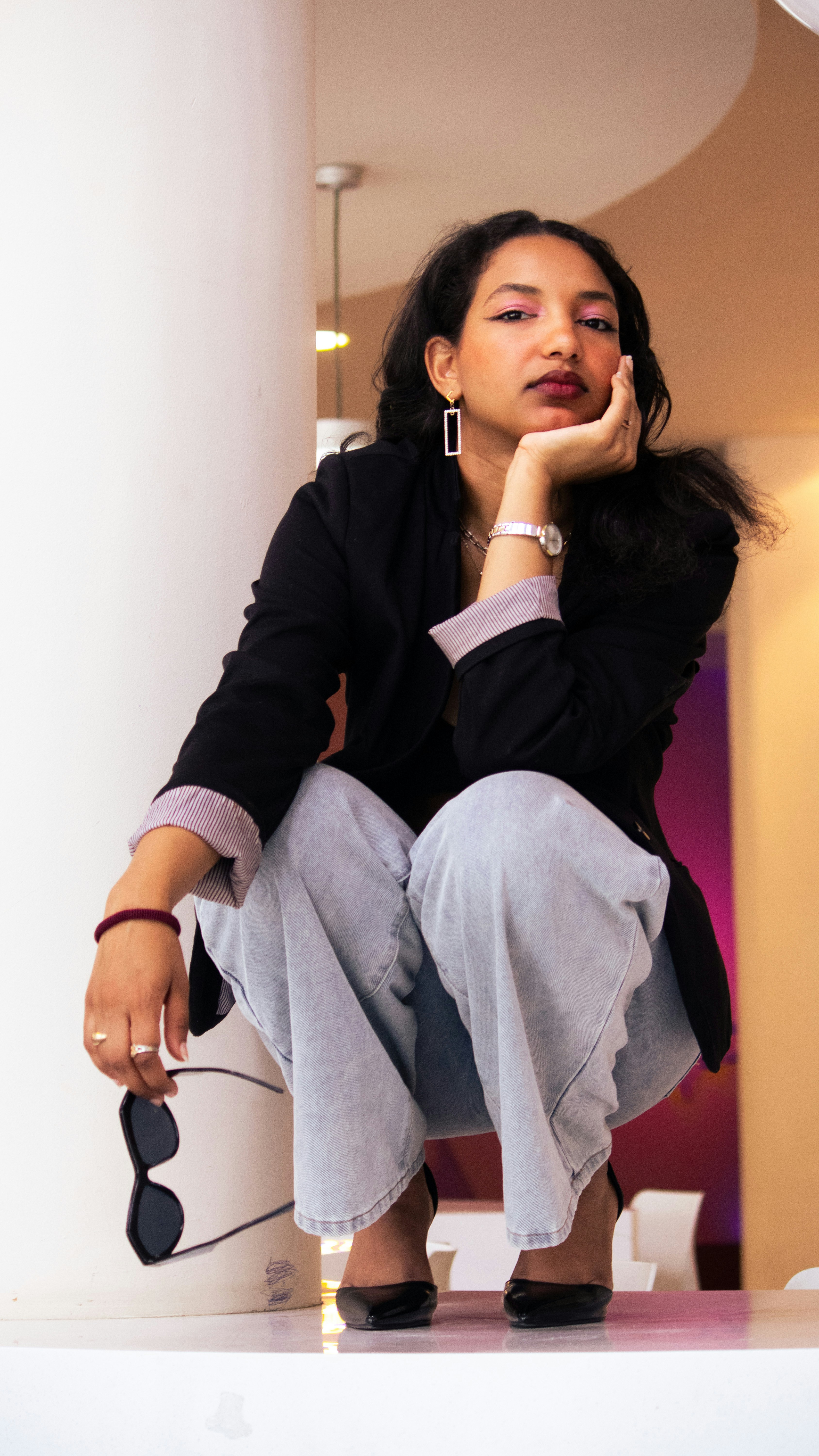 A stylish individual crouches gracefully on a ledge, holding sunglasses and exuding confidence in a modern urban setting.