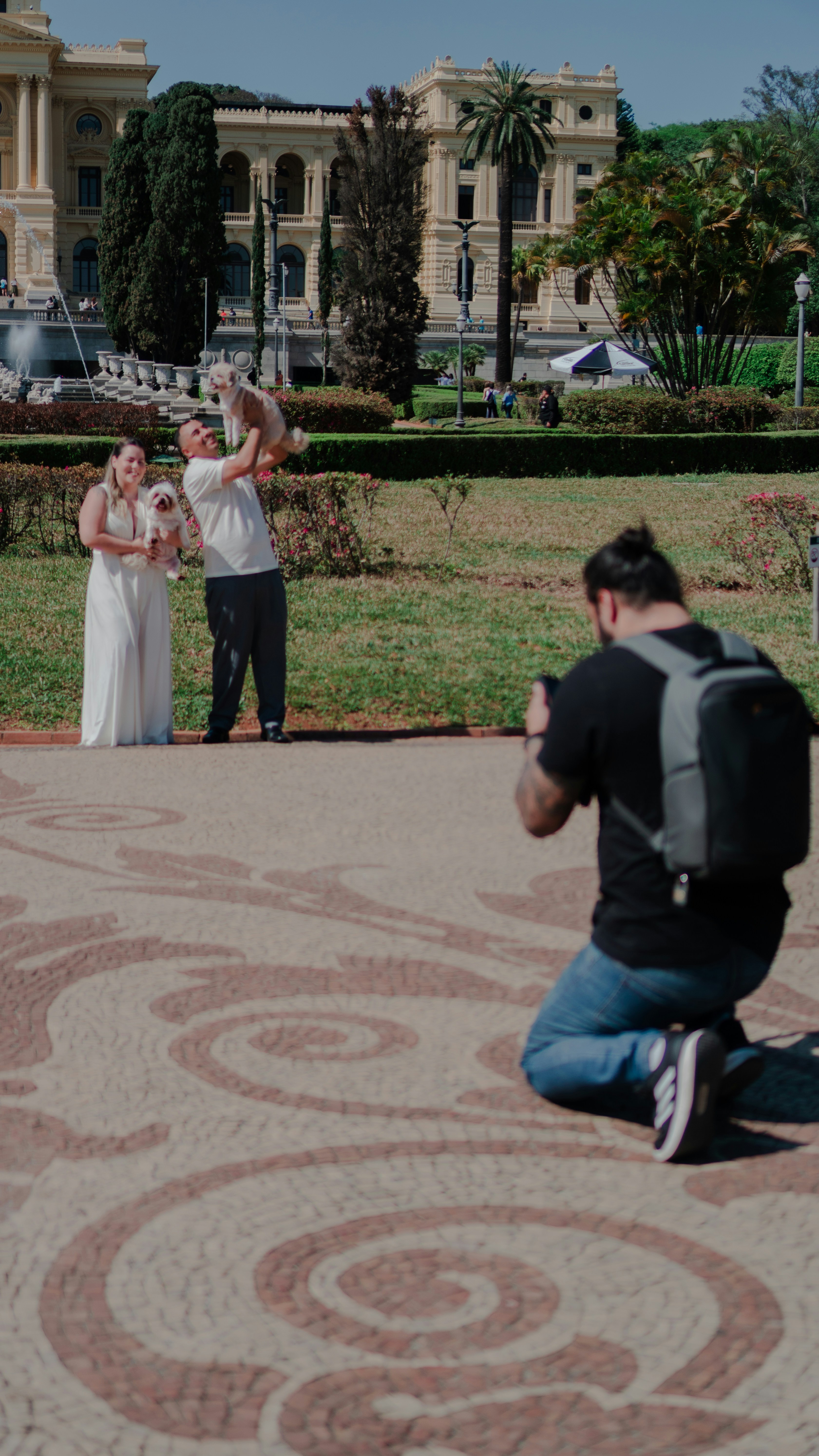 Couple in wedding attire holding their dog, posing for a photographer in a lush garden setting with an ornate building in the background.
