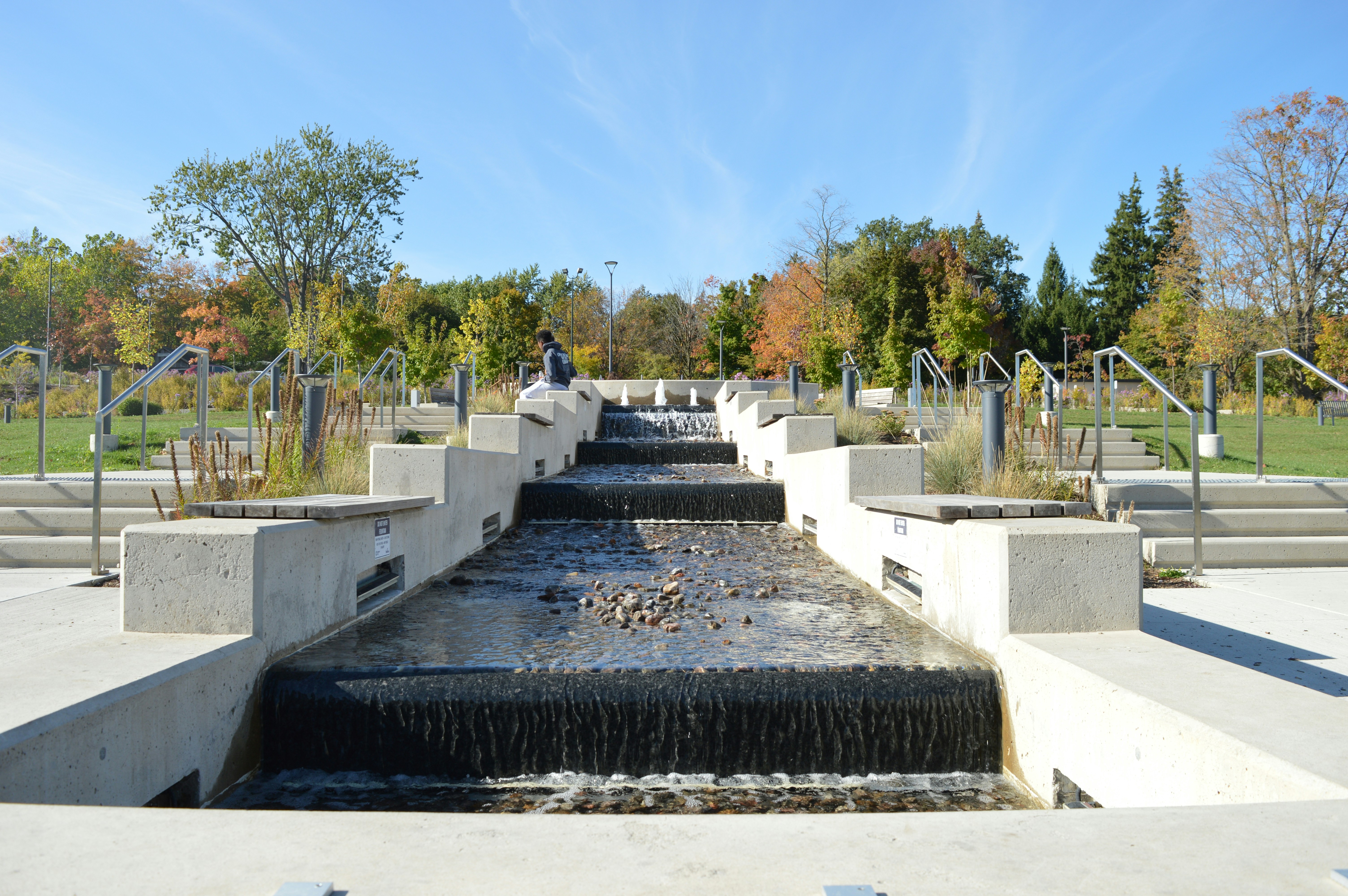 A modern water feature cascades down a series of stone steps, surrounded by greenery and autumn foliage. A solitary figure enjoys the peaceful scene.