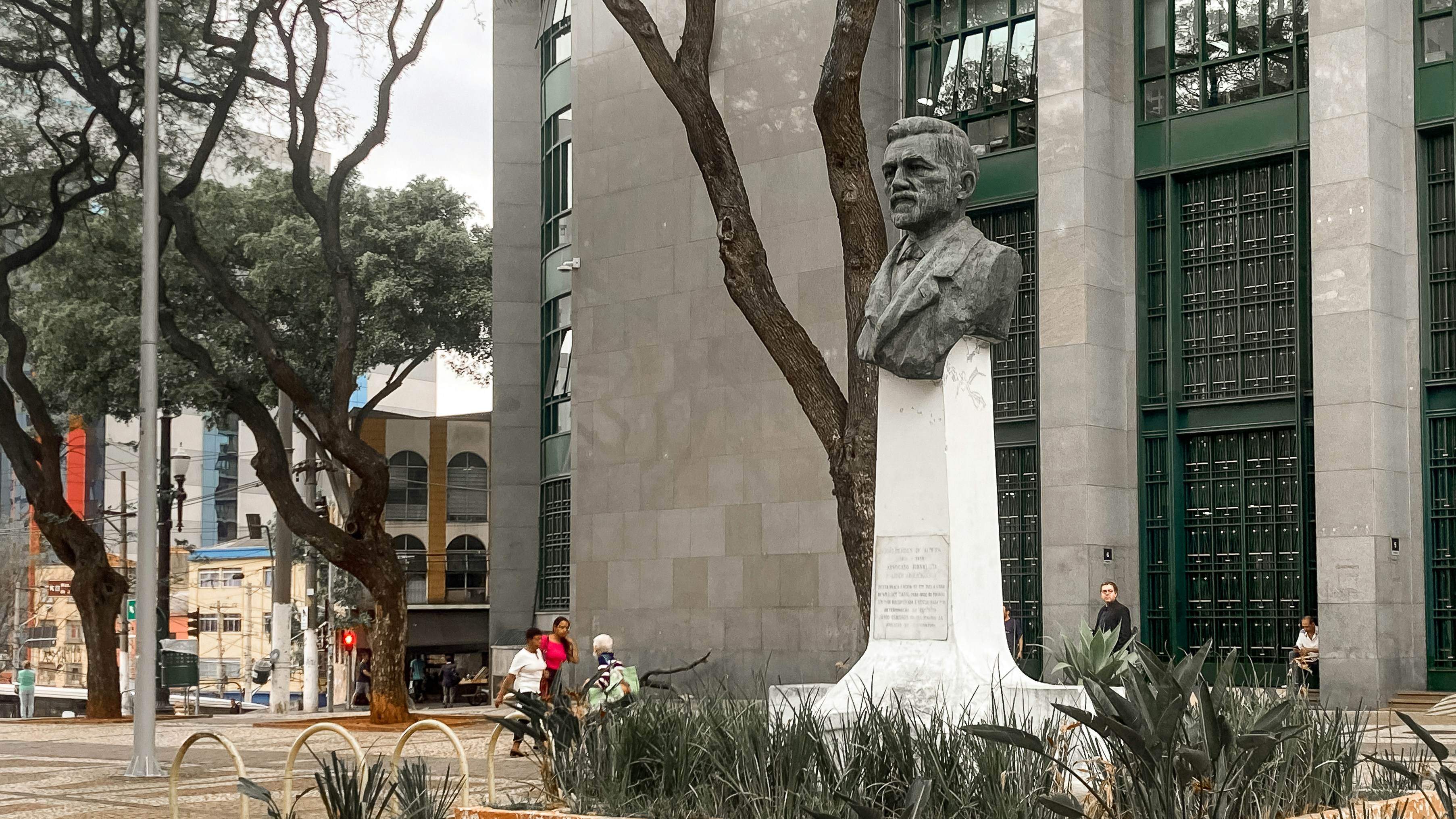 João Mendes de Almeida Júnior was a lawyer, journalist, and jurist born on March 30, 1856, in the city of São Paulo, Brazil. He became one of the most important jurists in Latin America, recognized for systematizing the theory of indigenato, thus contributing to guarantee rights for indigenous peoples. His career was also marked by being an excellent law professor at the Faculty of Law of Largo de São Francisco and concluding his trajectory as a Minister of the Federal Supreme Court. In 1913, João Mendes was honored with a bust sculpted by William Zadig, installed in the square in front of the São Paulo city courthouse. | Bust statue in front of a modern building