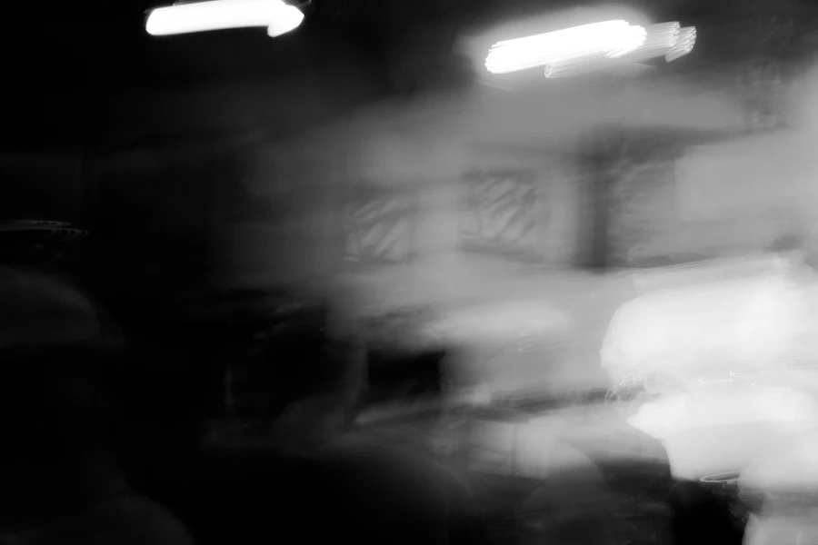 Black-and-white motion-blurred scene with bright overhead lights, a hazy stage area, and shadowy figures in the foreground. The right side glows with a blown-out subject or light source.