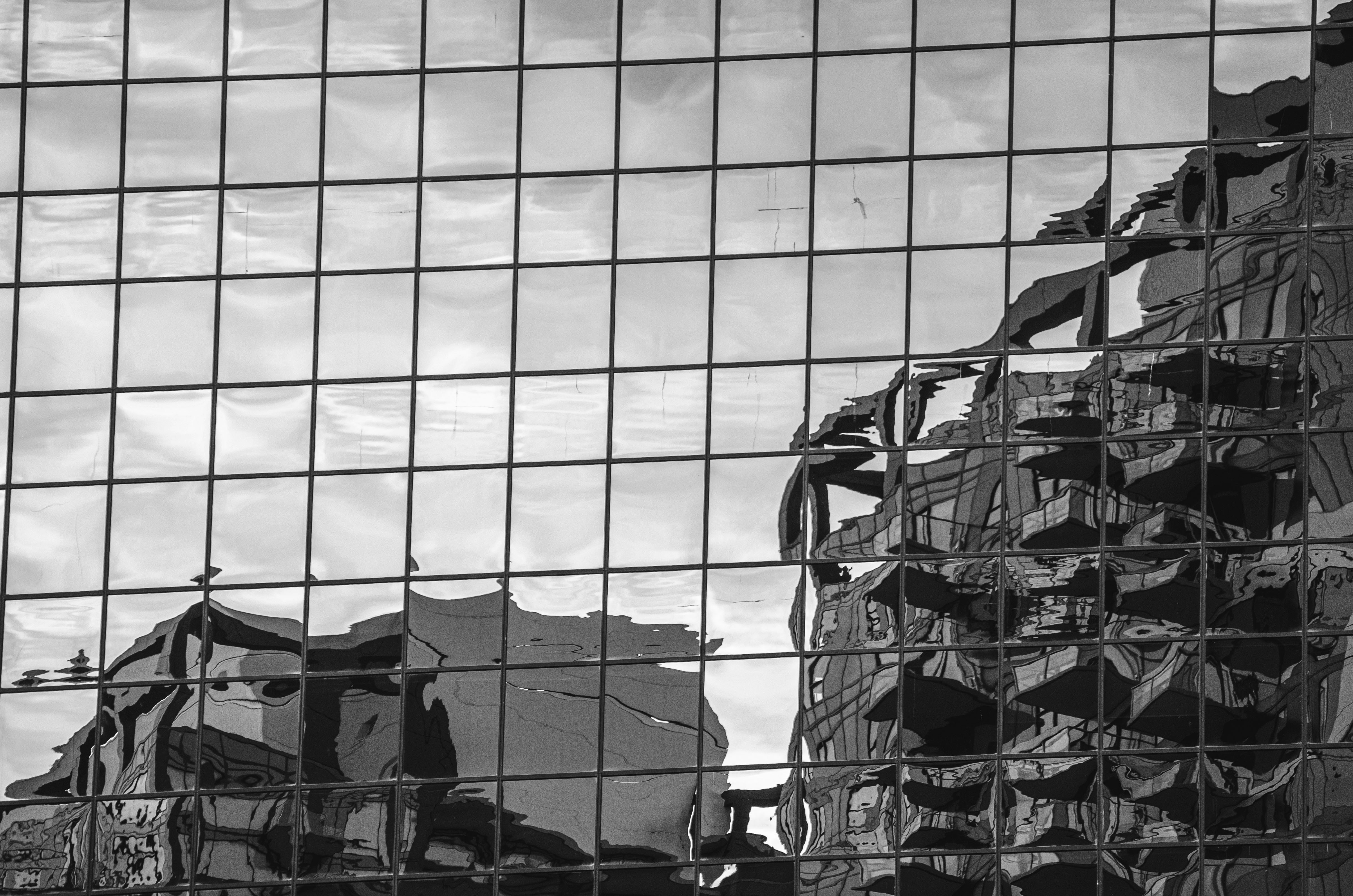 A black-and-white reflection of city buildings on a glass facade. The distorted reflections merge architectural elements with rippled glass, creating an abstract, layered composition. | Reflection of buildings in a grid window