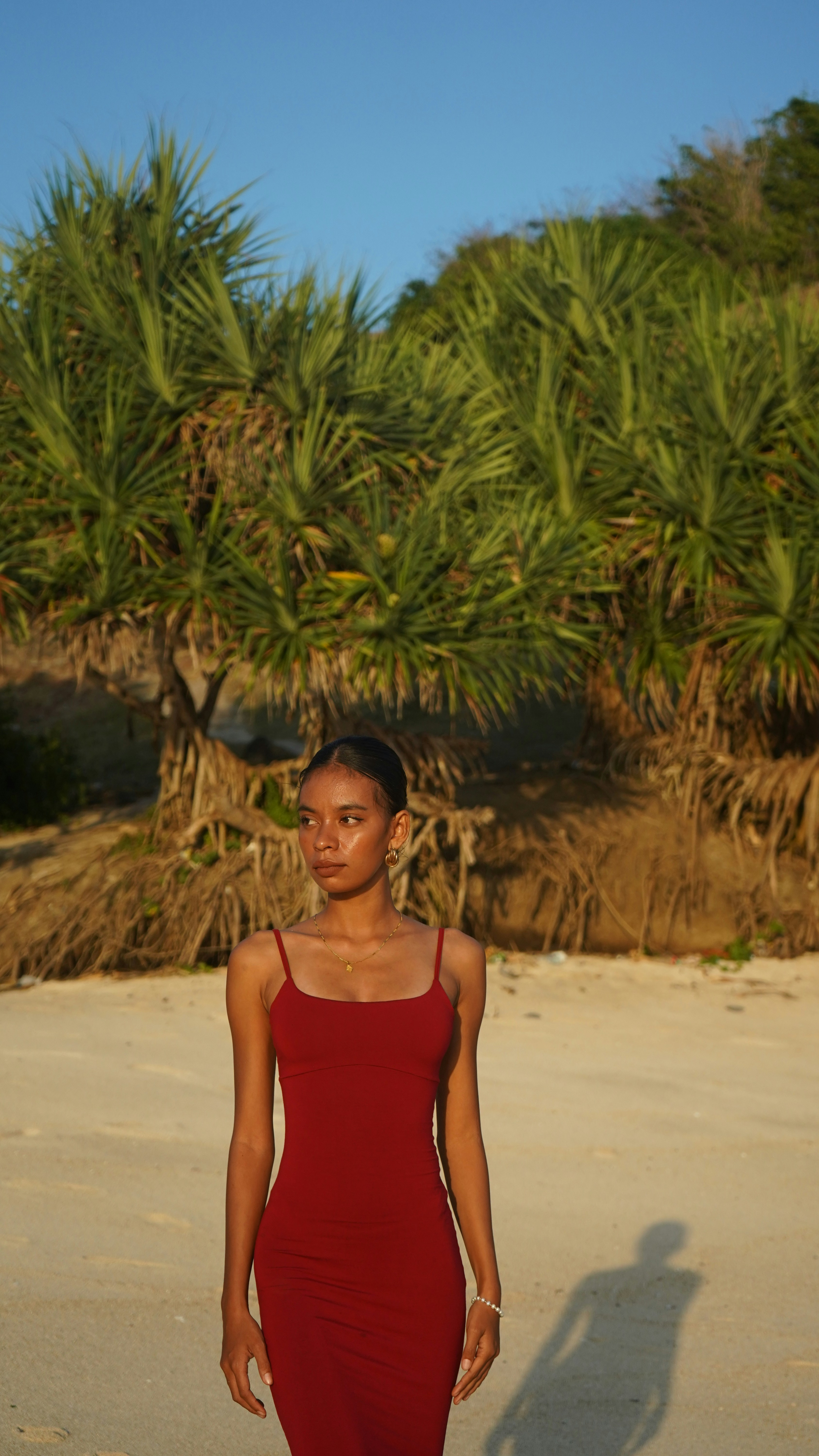 A woman in a fitted red dress stands on a sandy beach, framed by lush tropical foliage and a clear blue sky.