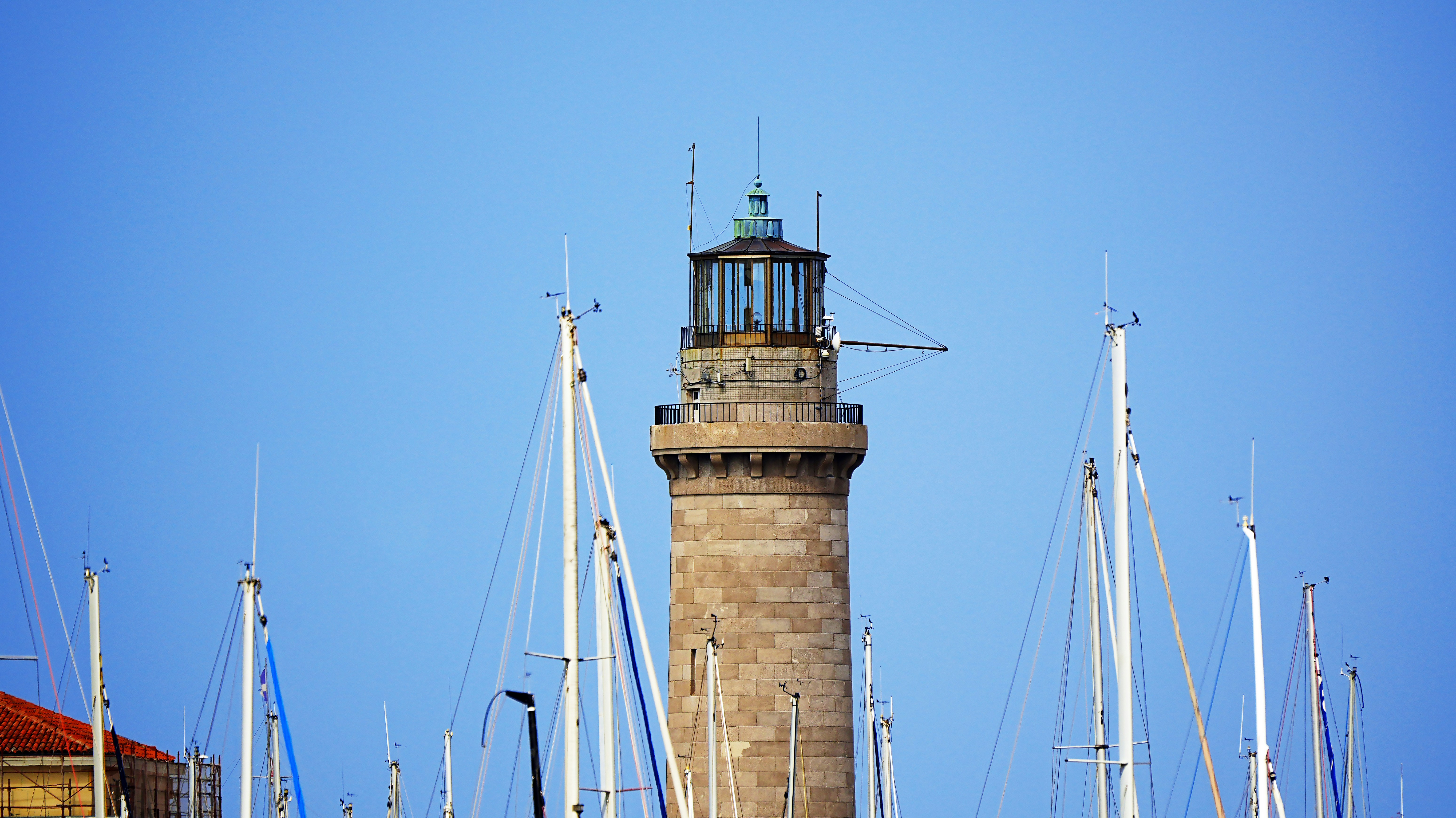 Stone lighthouse surrounded by sailboat masts against blue sky