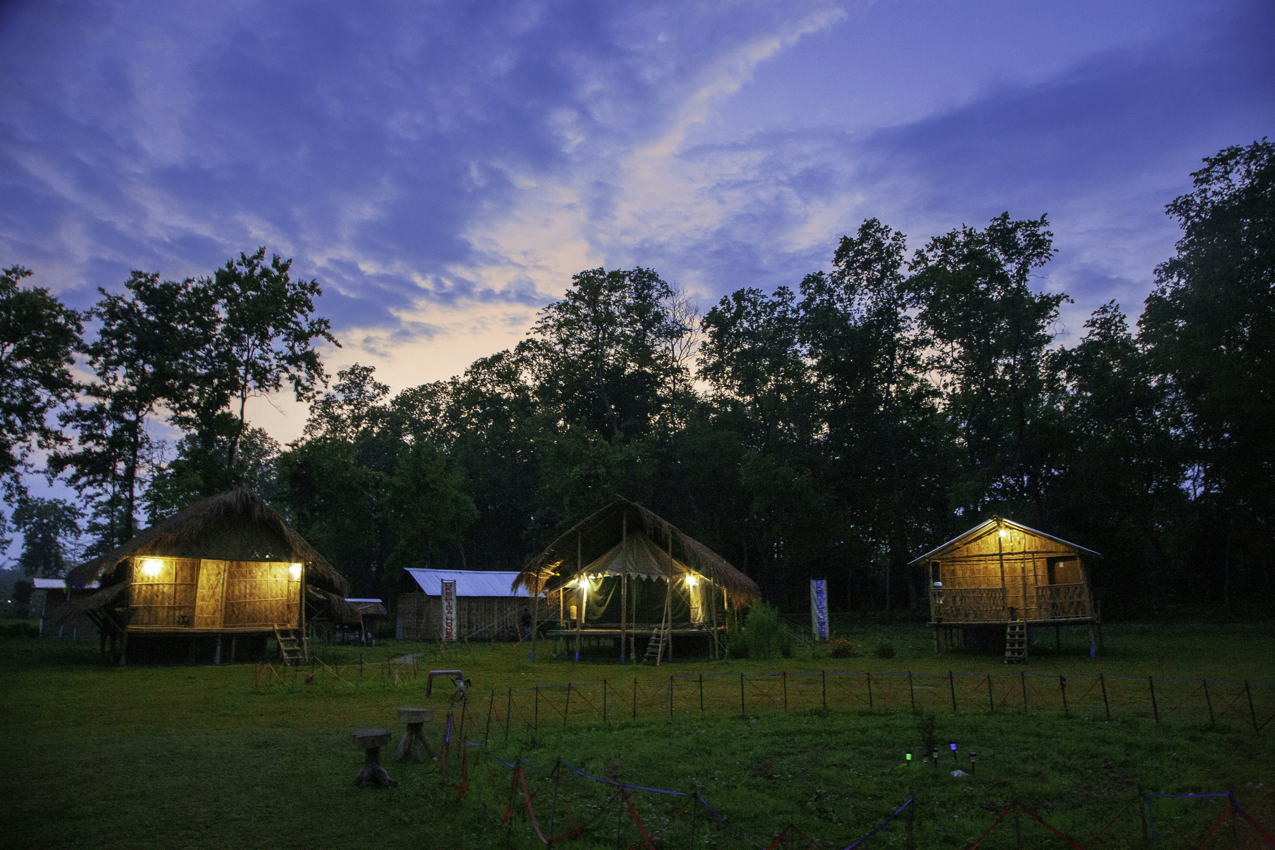 A beautiful Homestay. | Three huts illuminated at dusk in a wooded area.