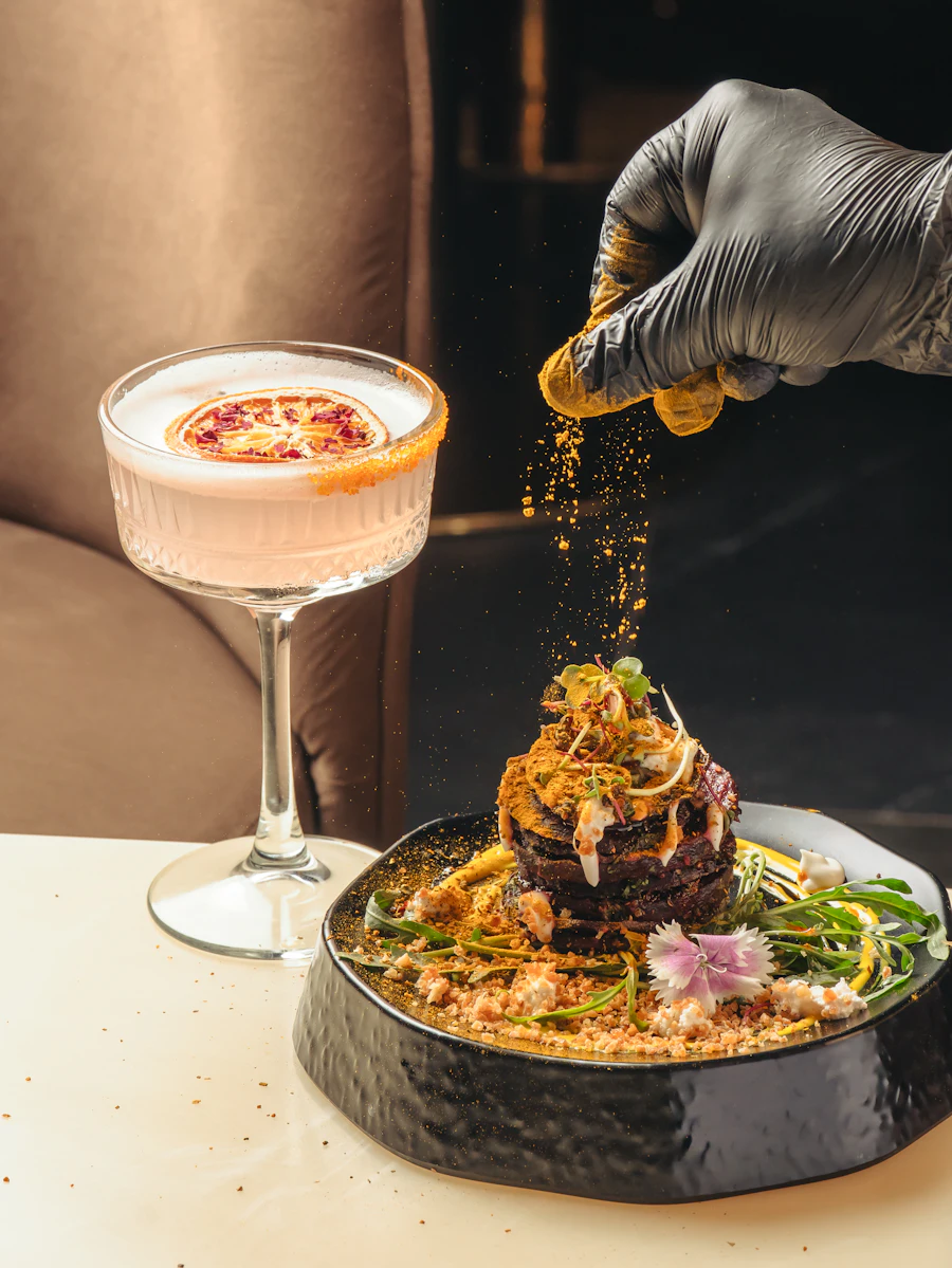 A gloved hand sprinkles powdered garnish over a stacked plated dessert beside a coupe glass filled with a pale pink cocktail. Warm restaurant lighting highlights the textured ingredients and elegant presentation.