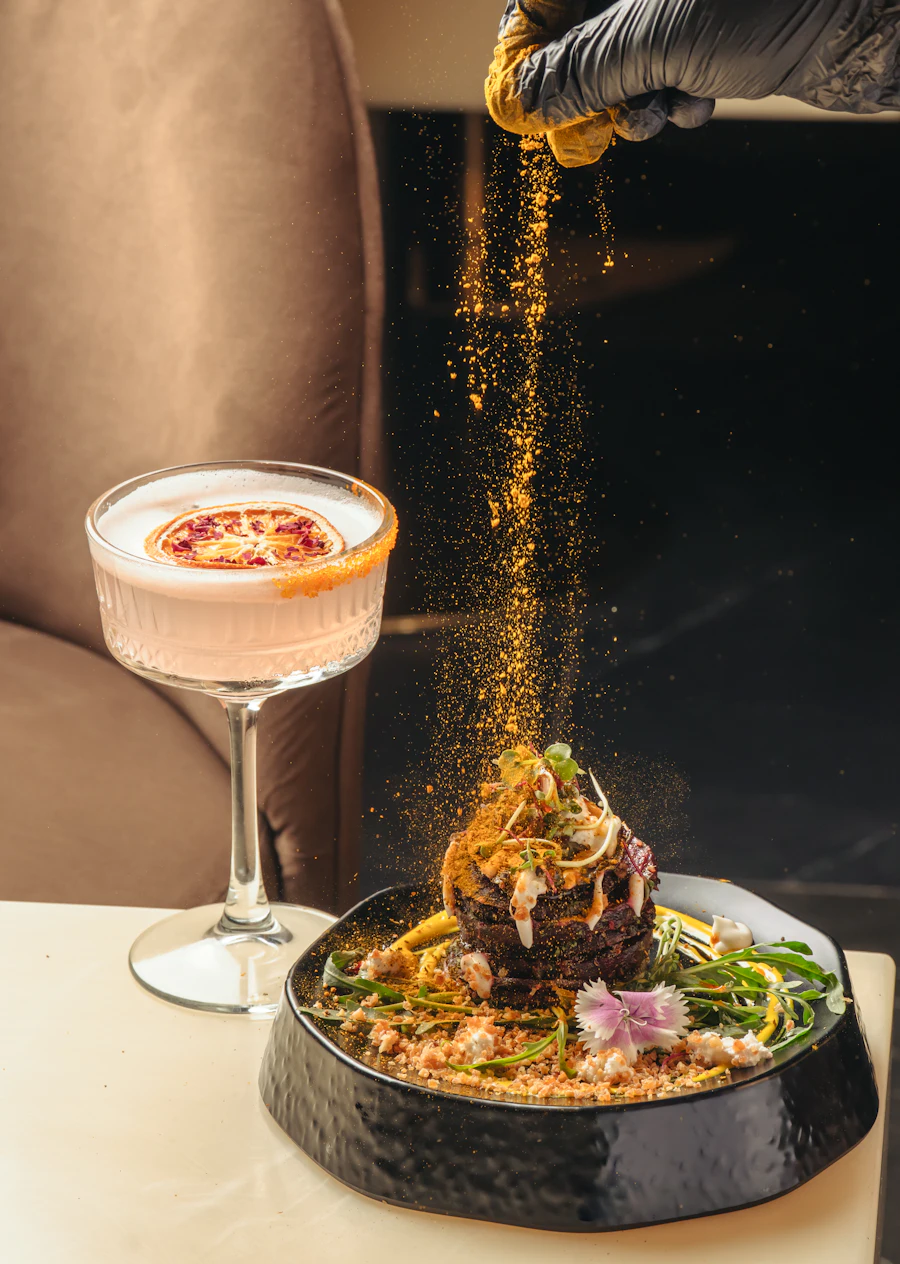 A plated gourmet dessert or savory dish is finished with a shower of golden powder beside a coupe cocktail topped with a dried citrus slice. The scene is styled on a table with dramatic warm lighting.