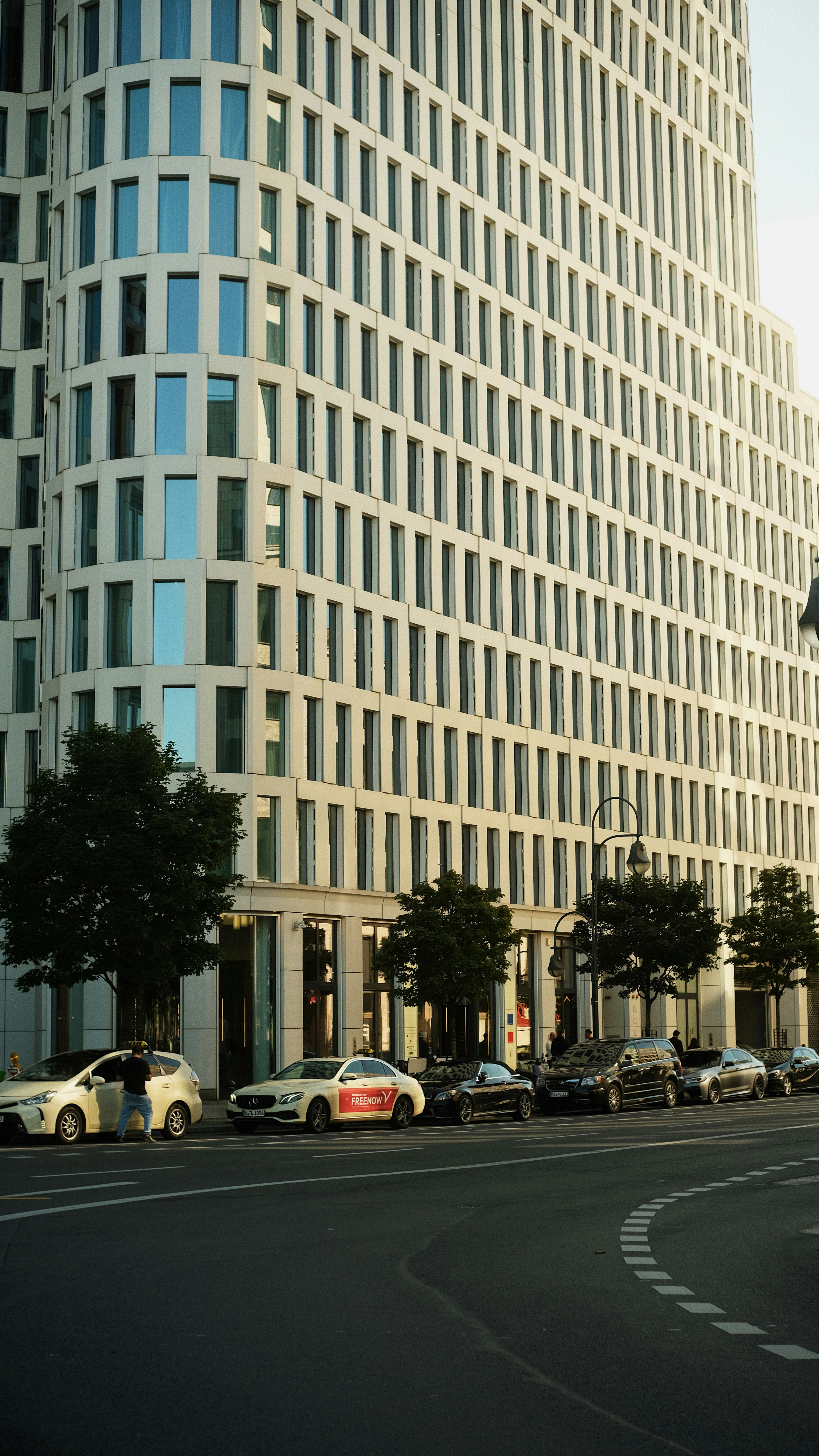 A contemporary building featuring a grid of reflective windows, framed by lush trees and parked cars along the street.