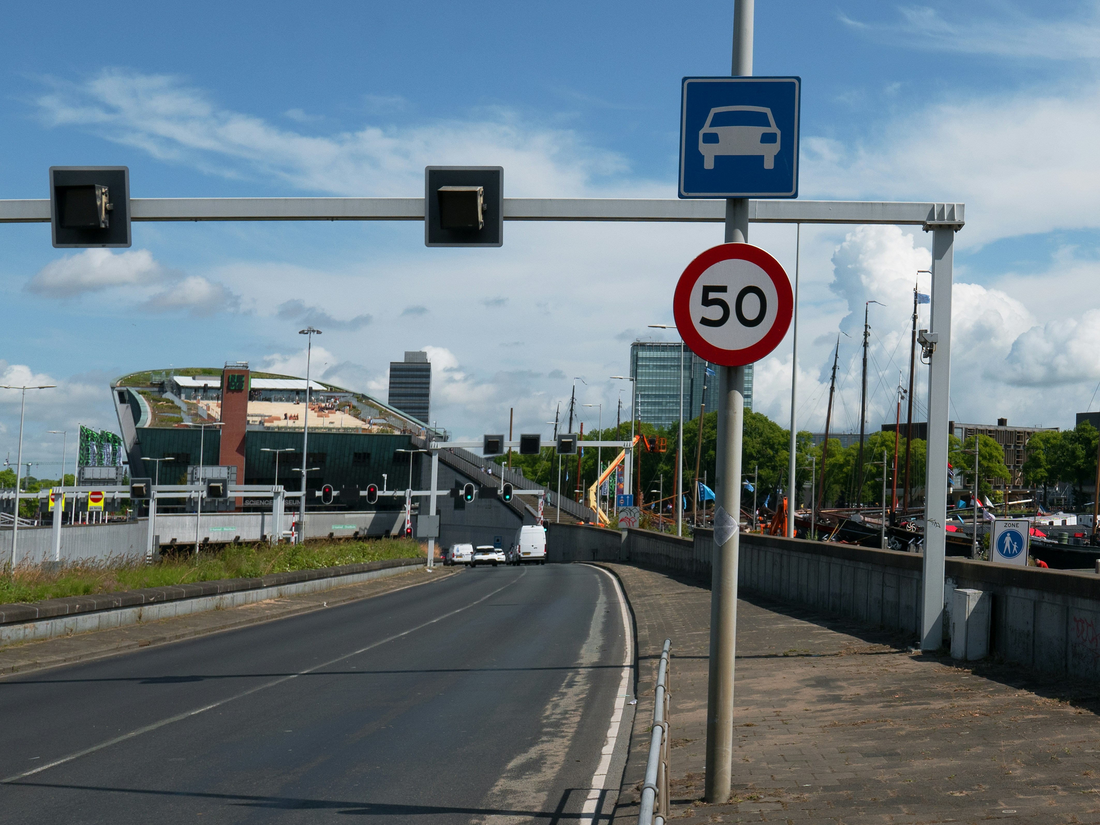 Ohoto of a view over the road going down to the IJ-tunnel under the IJ water in Amsterdam, with street lights and traffic signs above the road.. Free image under a blue sky. | Road with speed limit sign and traffic lights