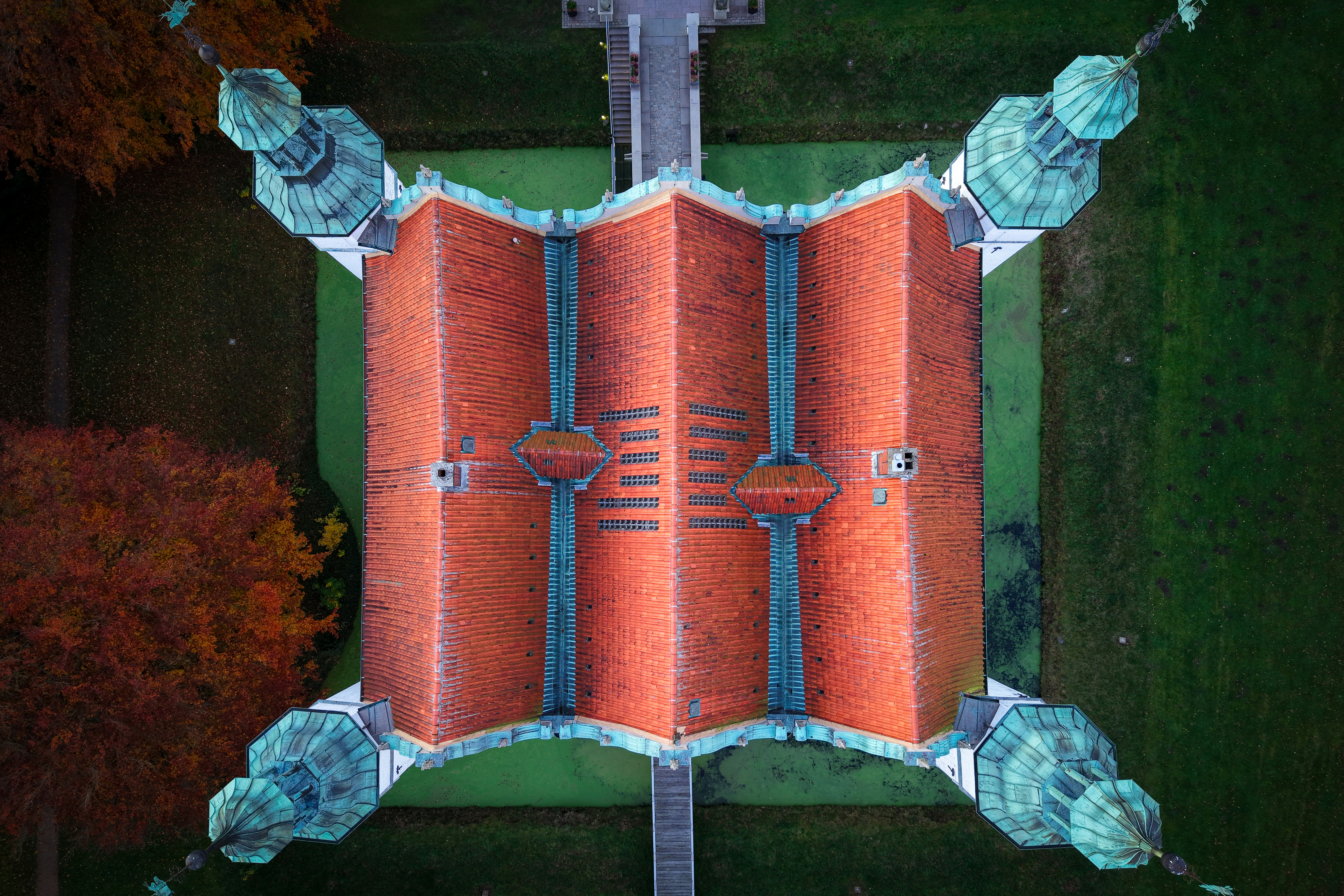 Perfect symmetry from above ✨ Captured with the DJI Air 3S, this top-down aerial shot of Ahrensburg Castle reveals the stunning geometry and copper-green towers of one of Northern Germany’s architectural gems. The rich contrast between the red rooftops, green surroundings, and autumn tones creates a painterly balance — where precision meets artistry. 🚁📸 More on https://droneography.work | Aerial view of a building with a red tiled roof.