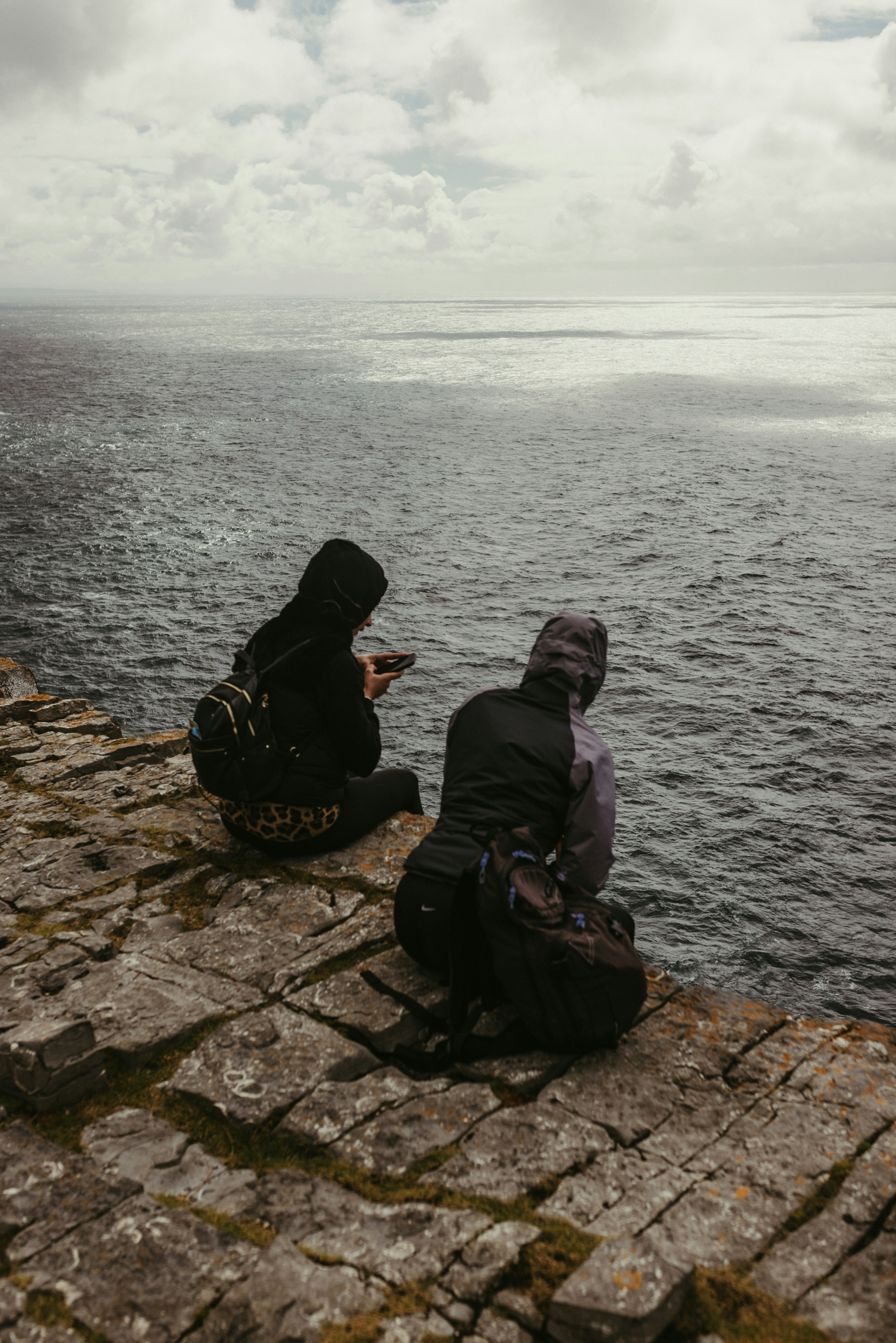 Two people sit on a cliff overlooking the ocean.