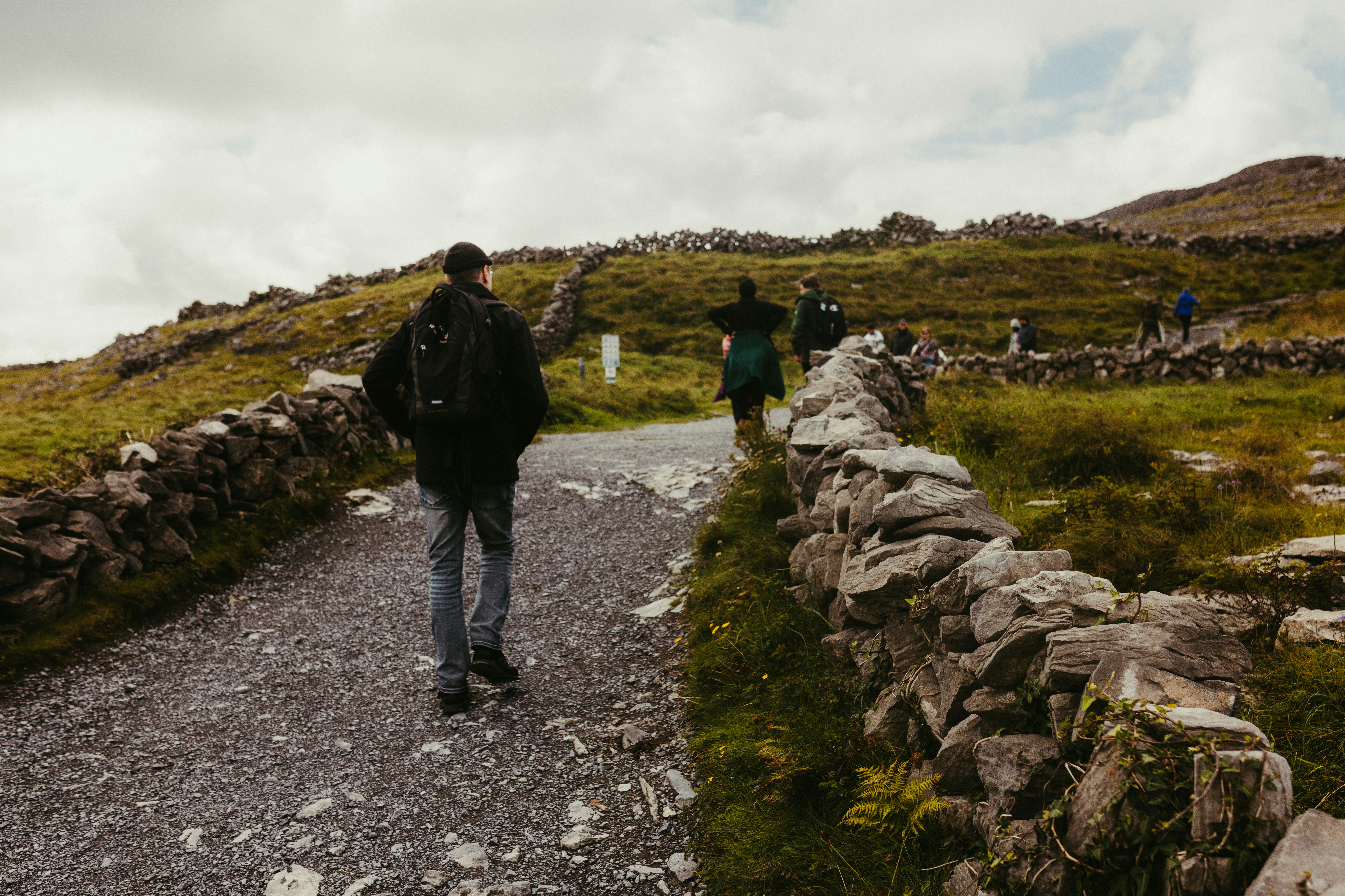 Hikers navigate a winding path lined with stone walls, surrounded by lush greenery and rolling hills.
