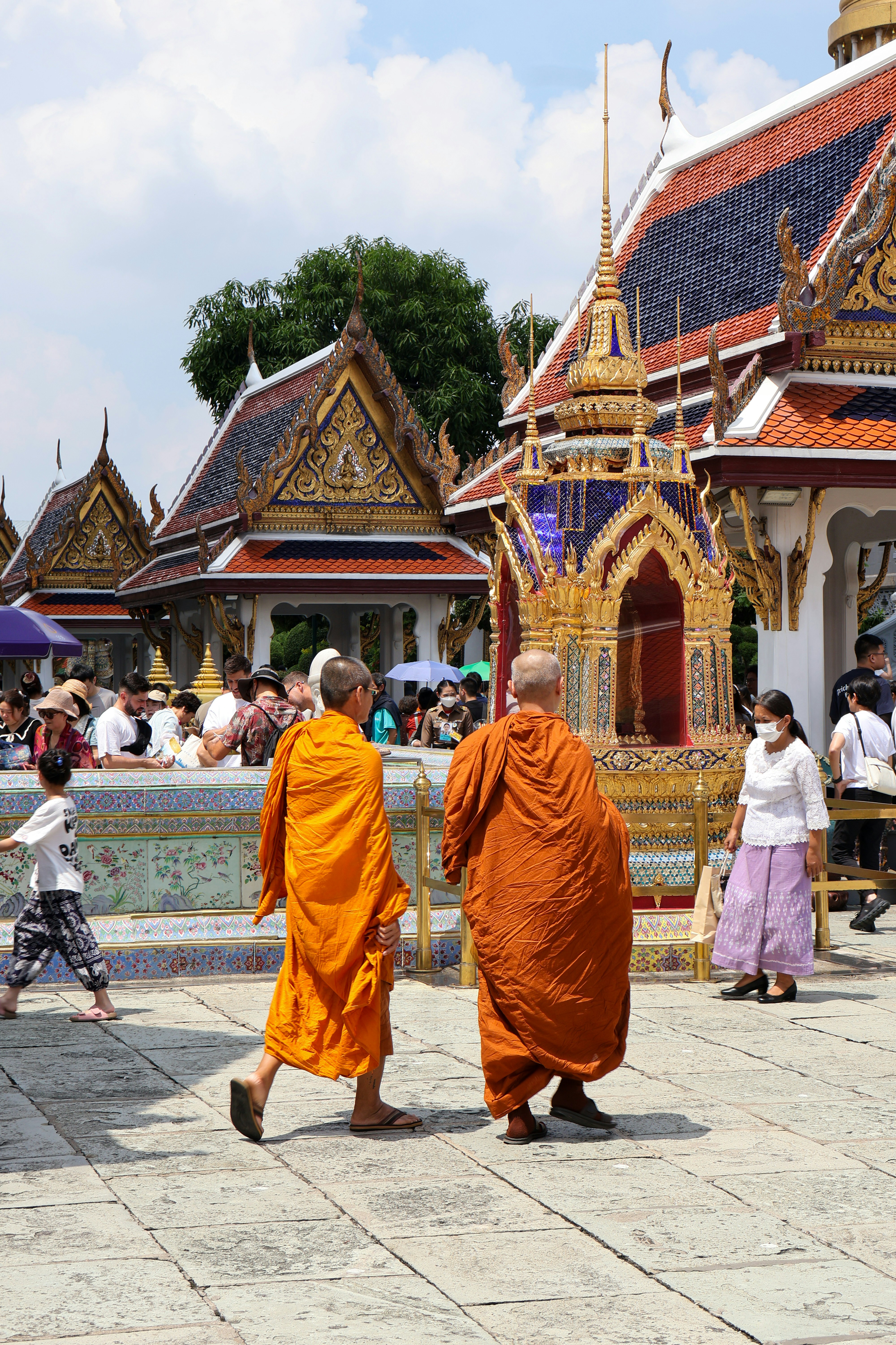 Grand Palace complex in Bangkok, Thailand, featuring Buddhist monks and visitors amidst the ornate architecture of a temple, likely Wat Phra Kaew (Temple of the Emerald Buddha). | Two monks walk past ornate temple buildings
