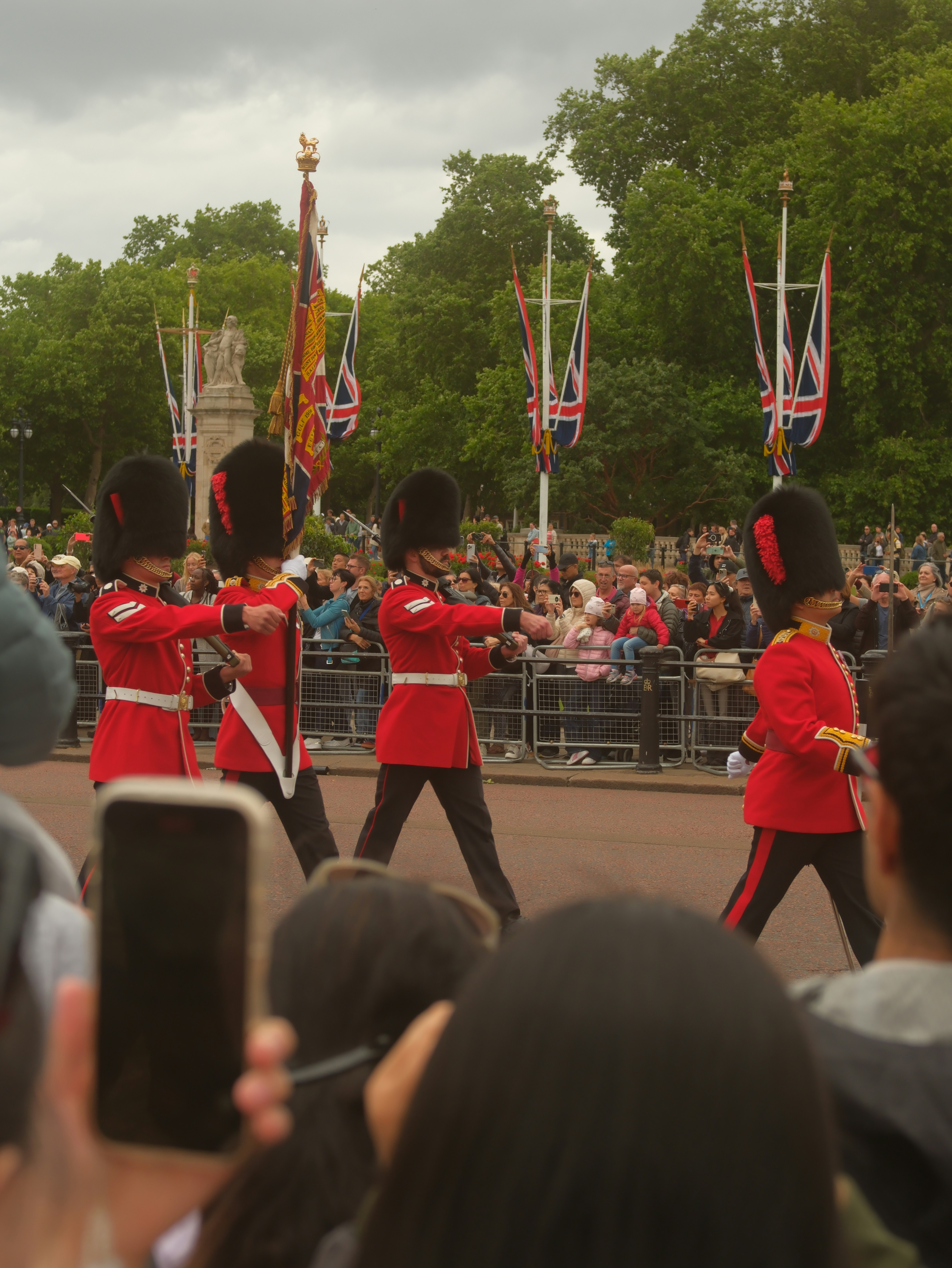 Ceremonial guards in traditional red uniforms march in front of a crowd, with Union Jack flags waving in the background.