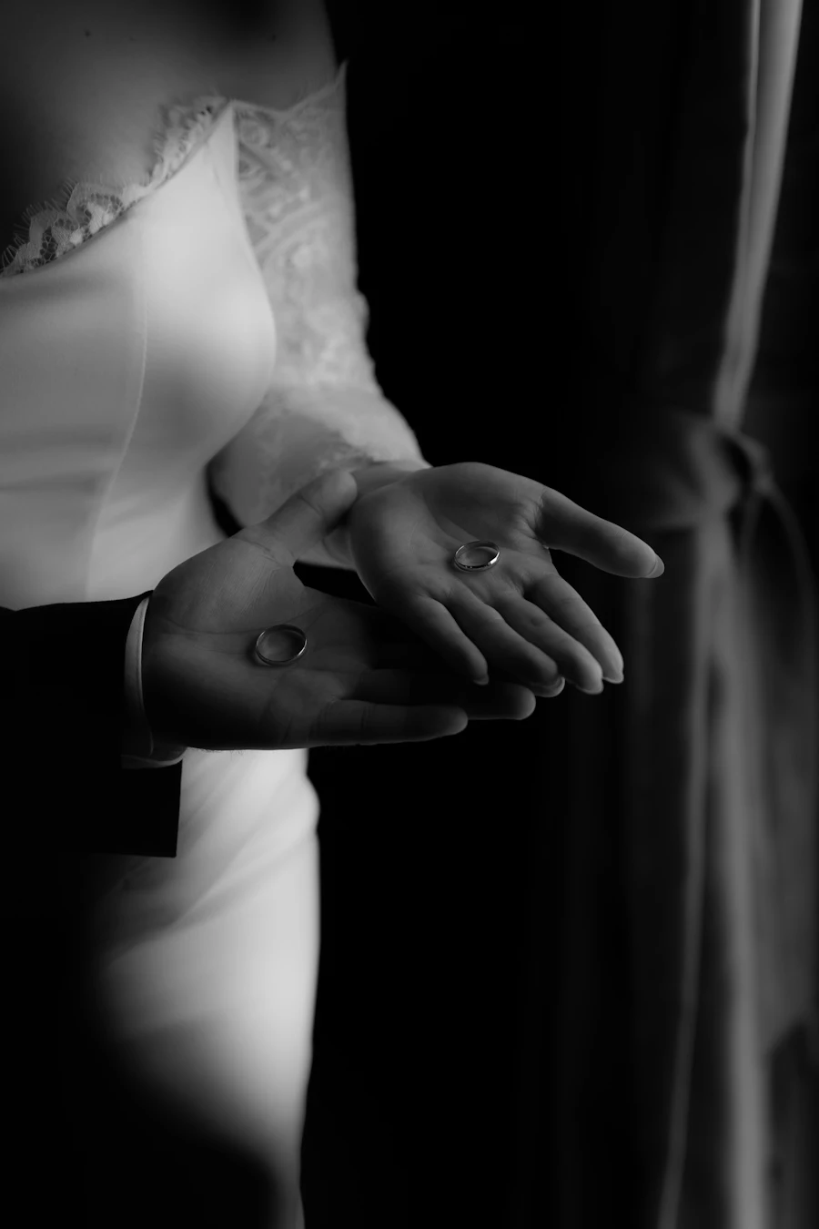 Black-and-white scene of a bride and groom holding wedding rings in open hands. The lace dress and suit sleeves frame the intimate moment.