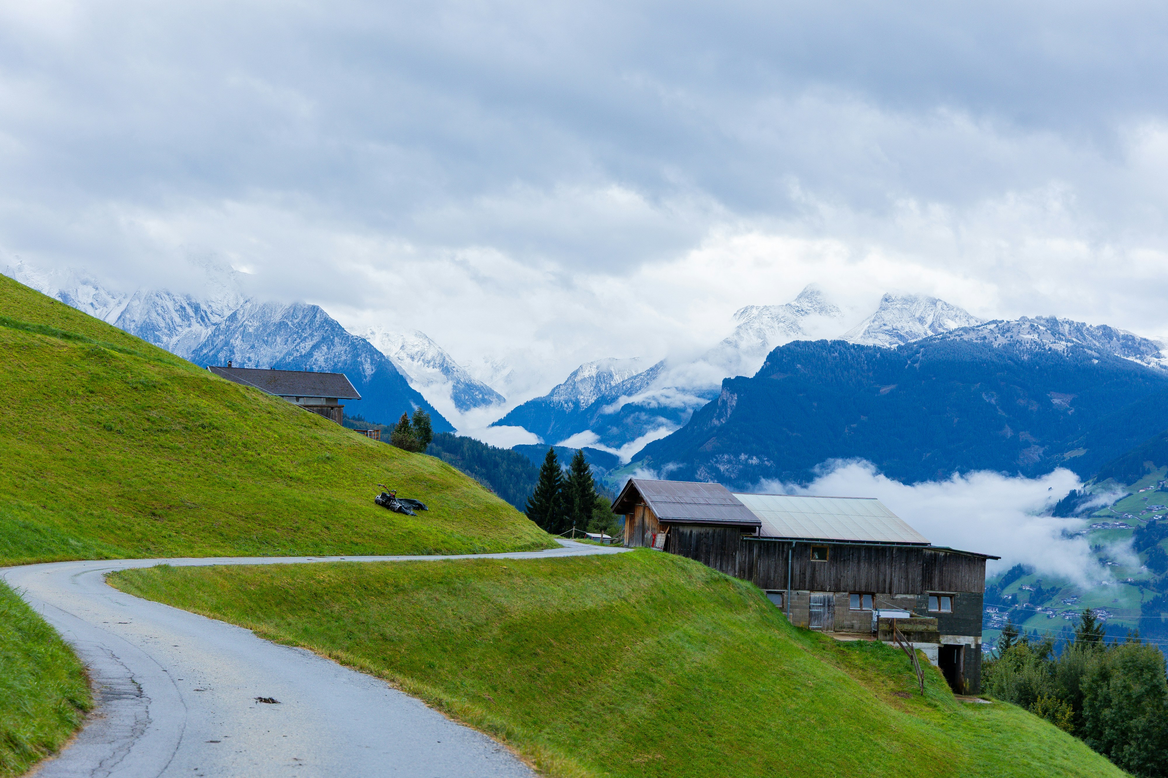 A rustic wooden cabin nestled on a verdant hillside, framed by majestic snow-capped mountains under a cloudy sky.