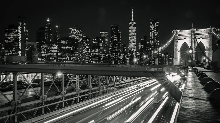 Black-and-white view from the Brooklyn Bridge with light trails leading toward the Manhattan skyline and the illuminated bridge tower.