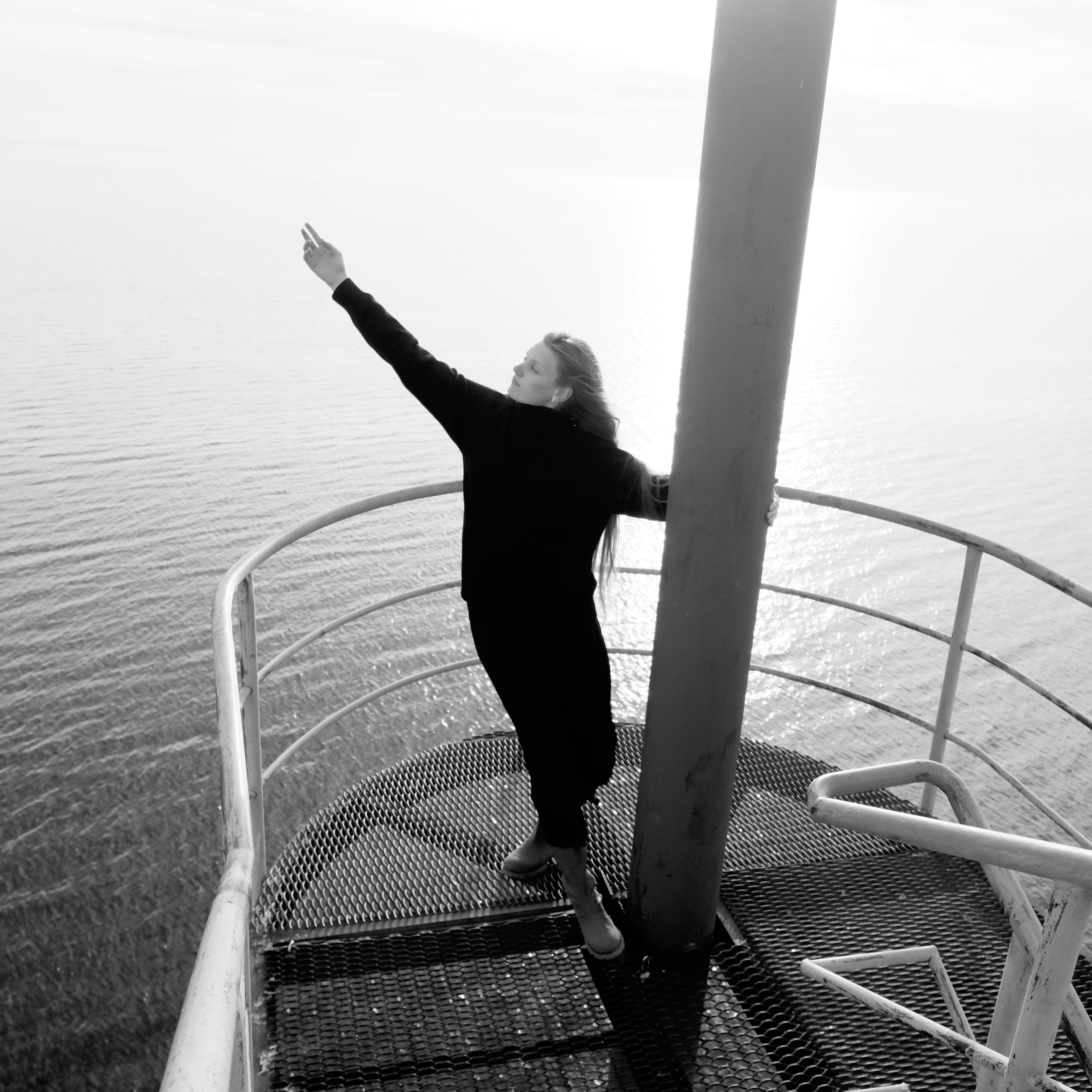 A figure in black clothing reaches out from a metal staircase on a pier, overlooking a calm sea at dawn. The scene is rendered in monochrome, emphasizing the contrast between the subject and the serene water.