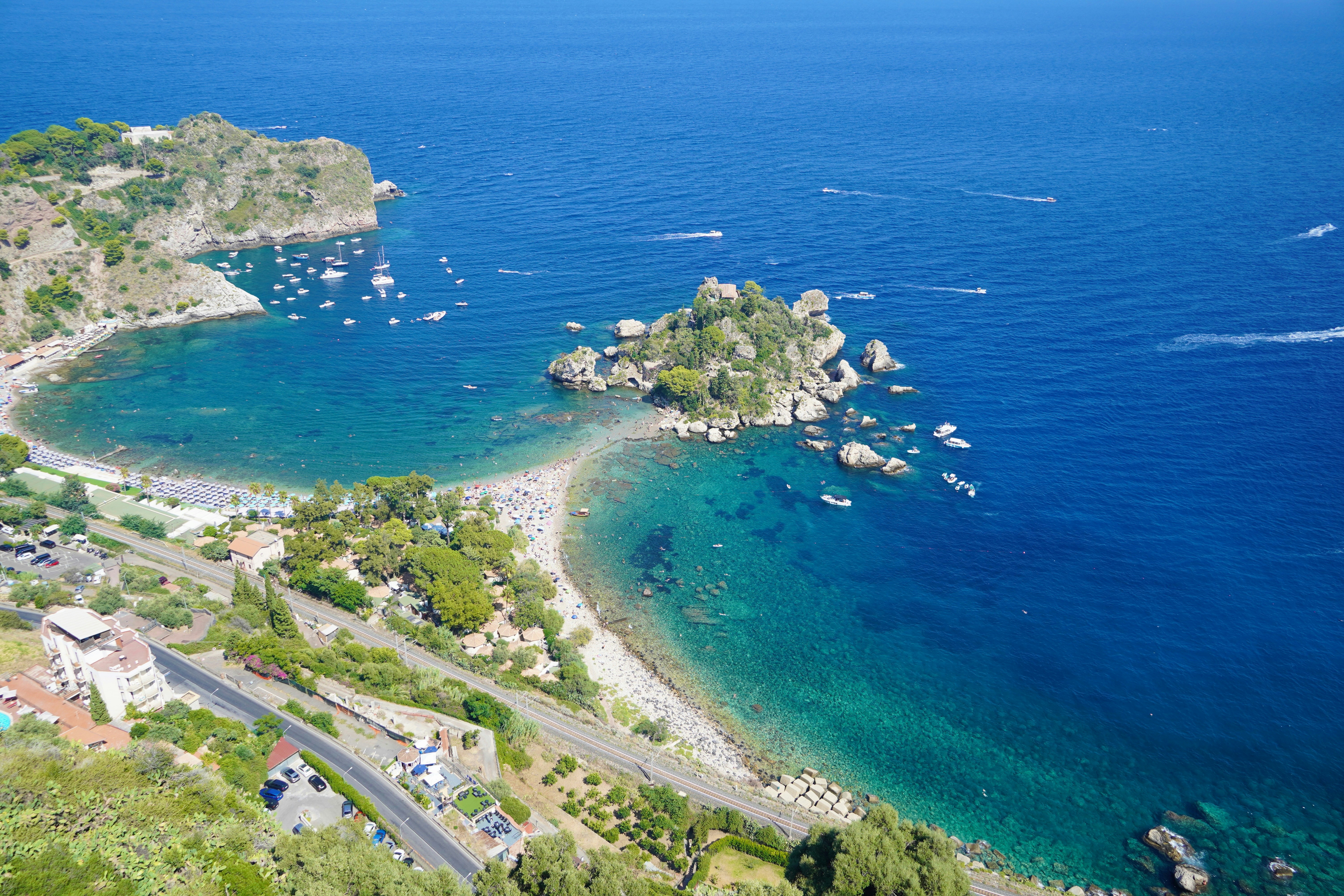 Aerial view of a picturesque coastline featuring a secluded beach, rocky islets, and vibrant turquoise waters. Boats dot the scene, enhancing the tranquil atmosphere.