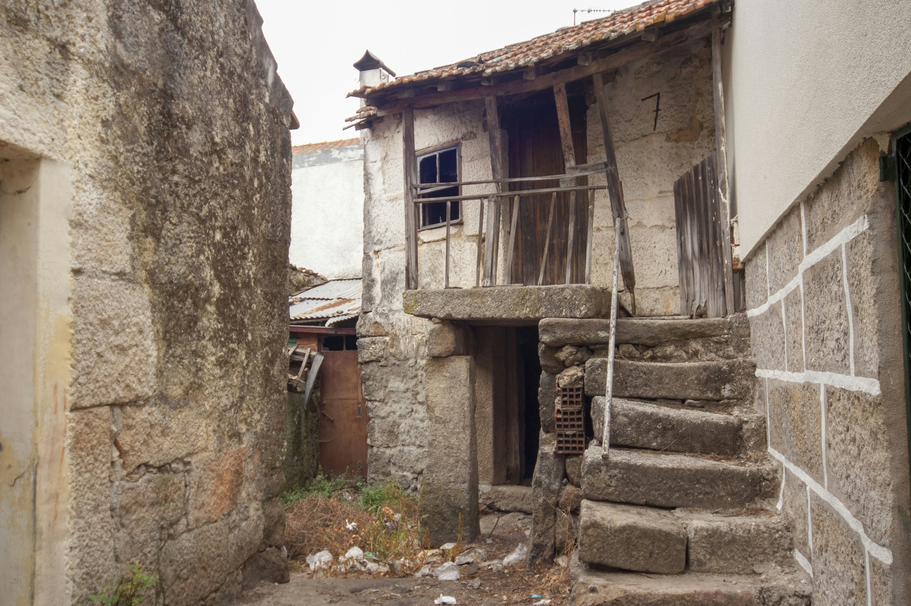 Abandoned rustic building with a wooden balcony and stone steps, nestled between weathered walls in a narrow alleyway.