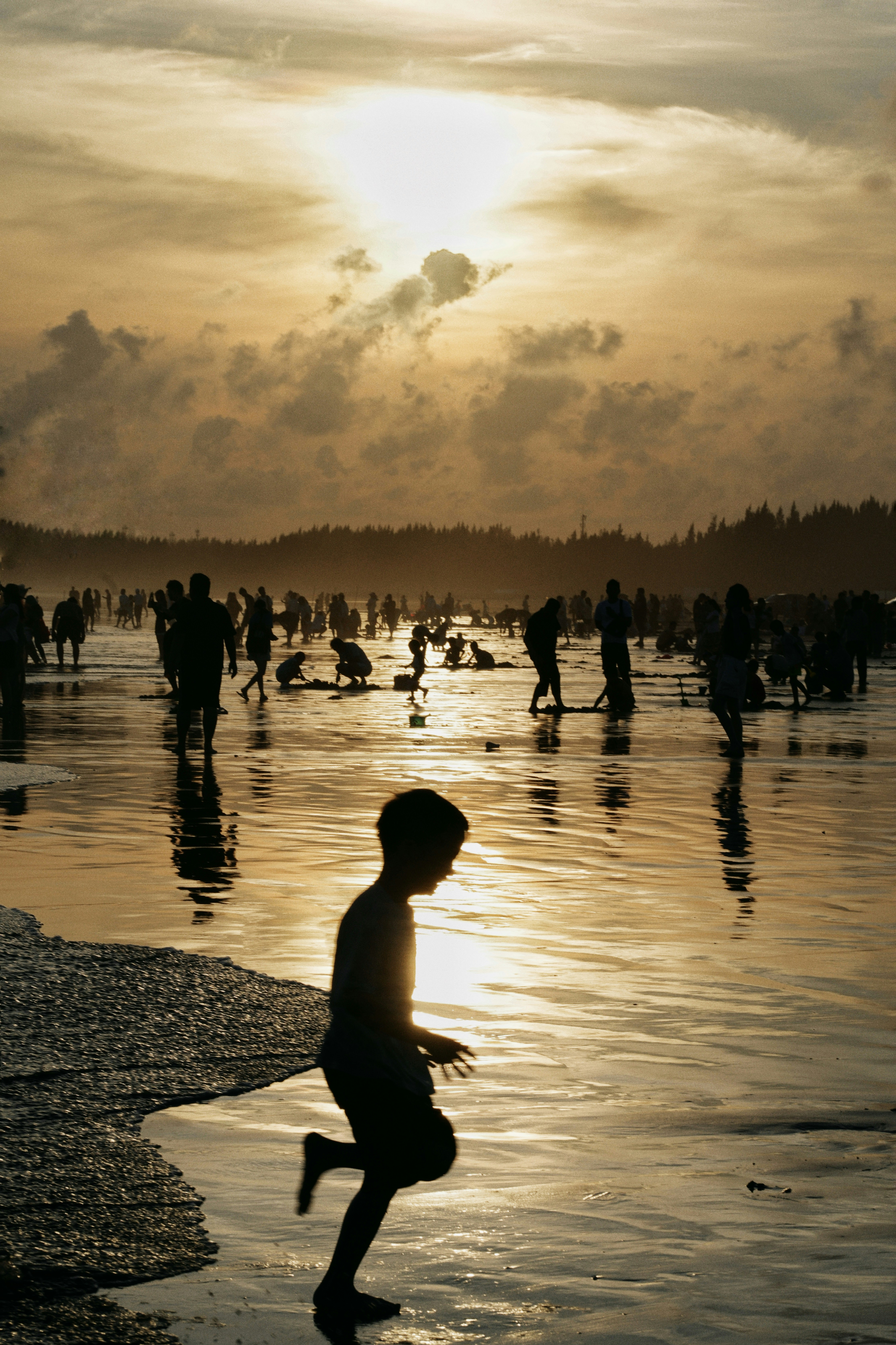 Silhouette of a child running along a reflective shoreline at sunset, surrounded by beachgoers enjoying the golden hour.