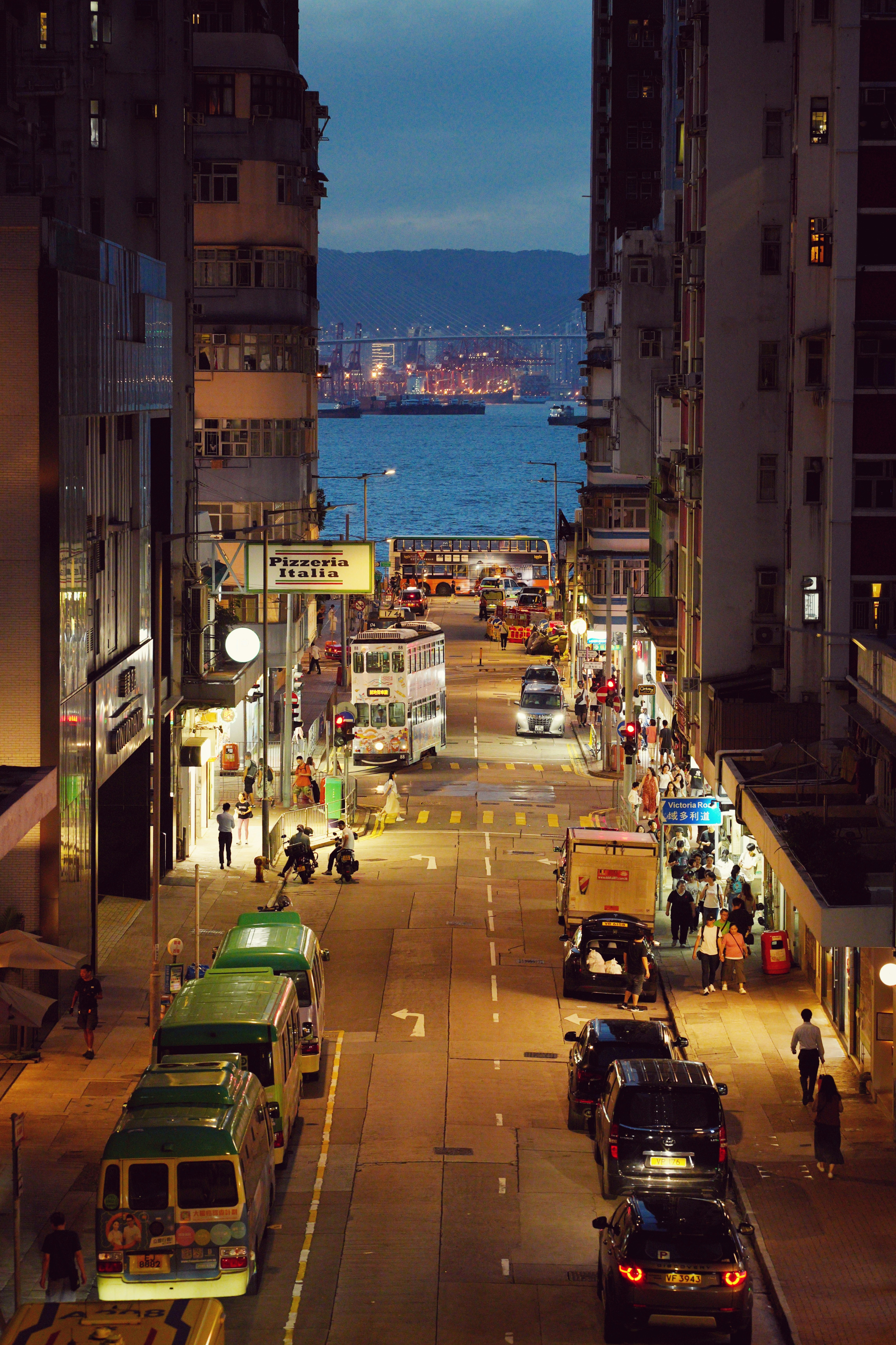 Vibrant street scene in a bustling city as twilight descends, showcasing vehicles, pedestrians, and city architecture. The distant harbor adds depth to the urban landscape.