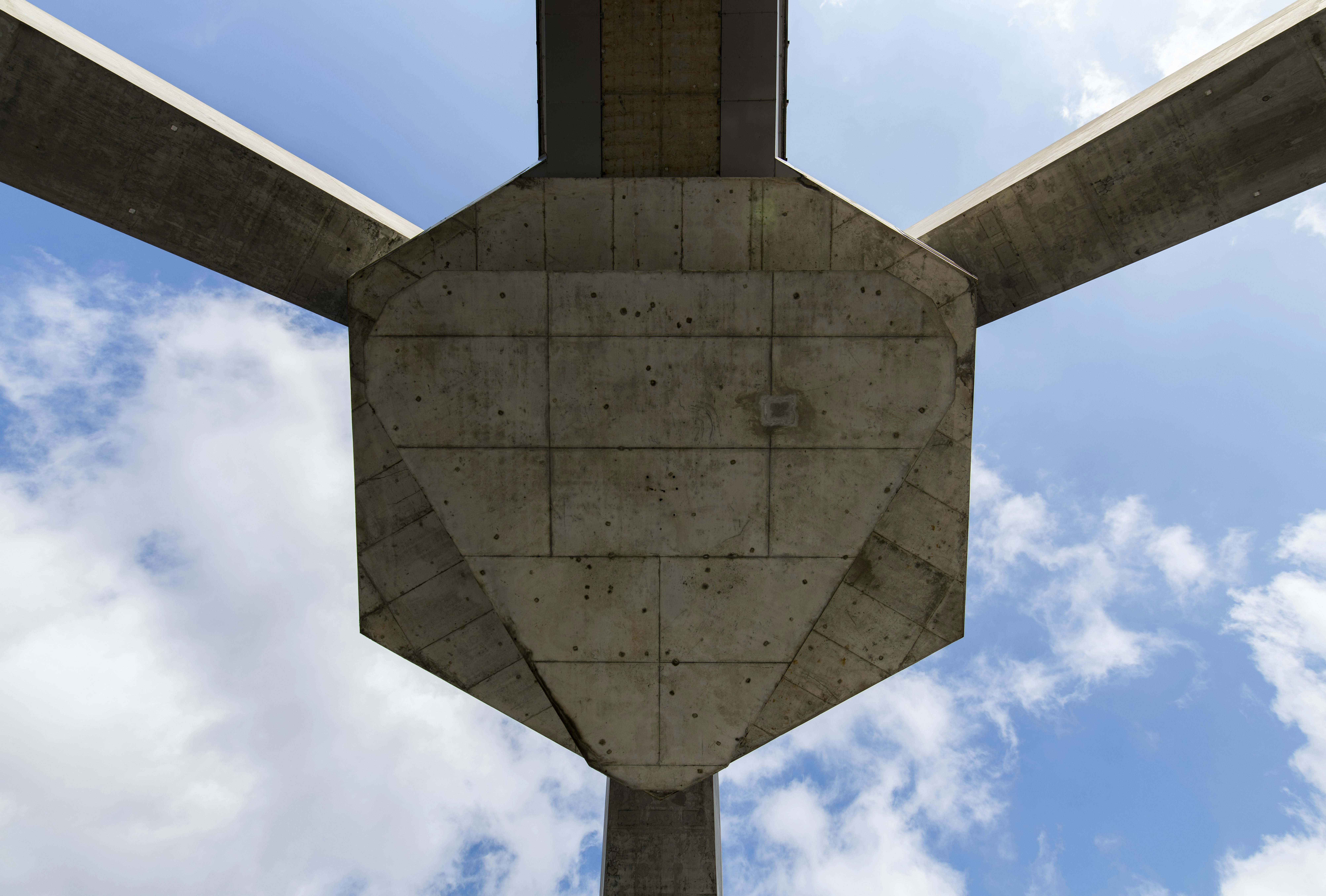 View of a concrete structure's underside, showcasing geometric patterns and textures against a backdrop of clouds.