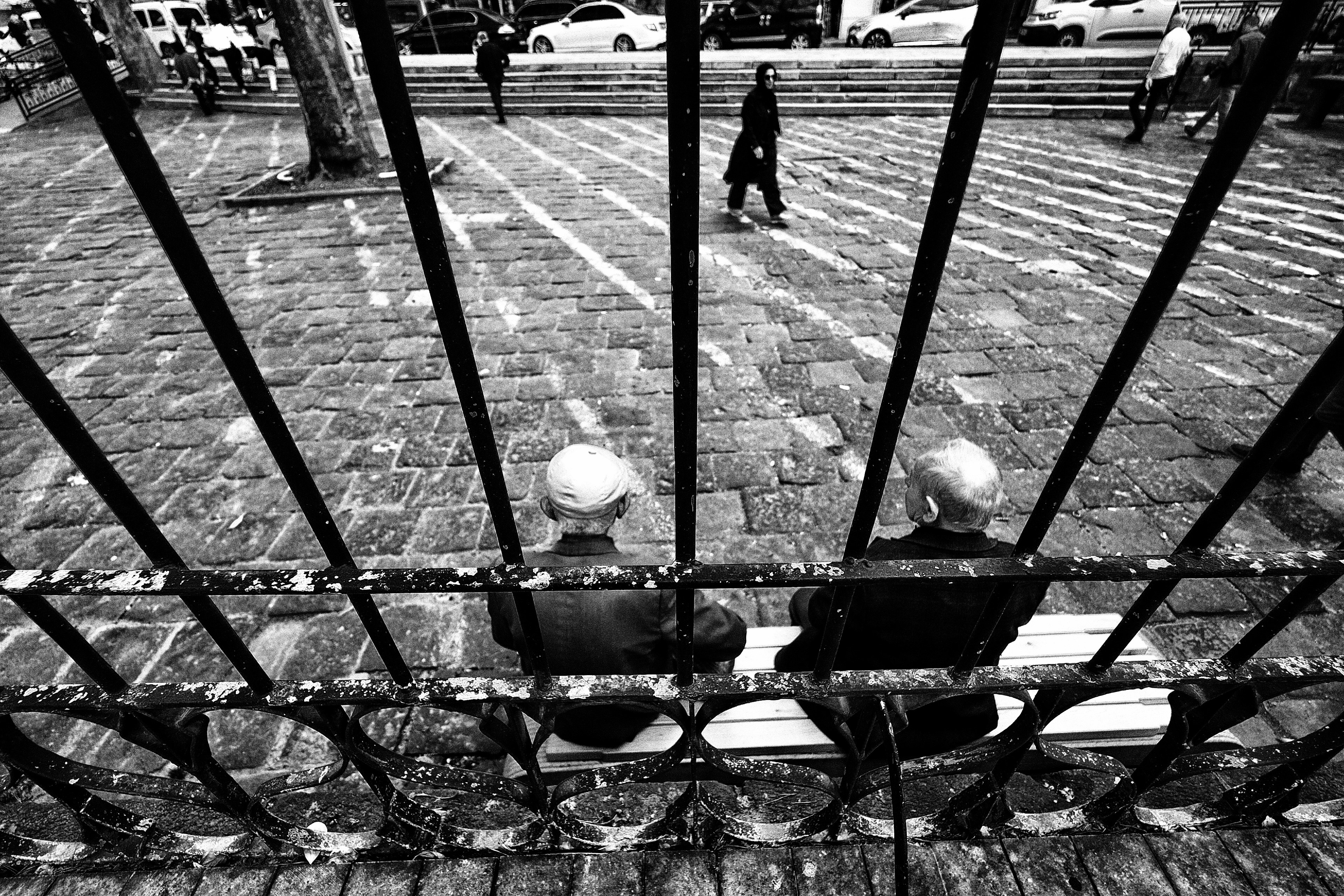 Two elders sit on a bench behind a fence.